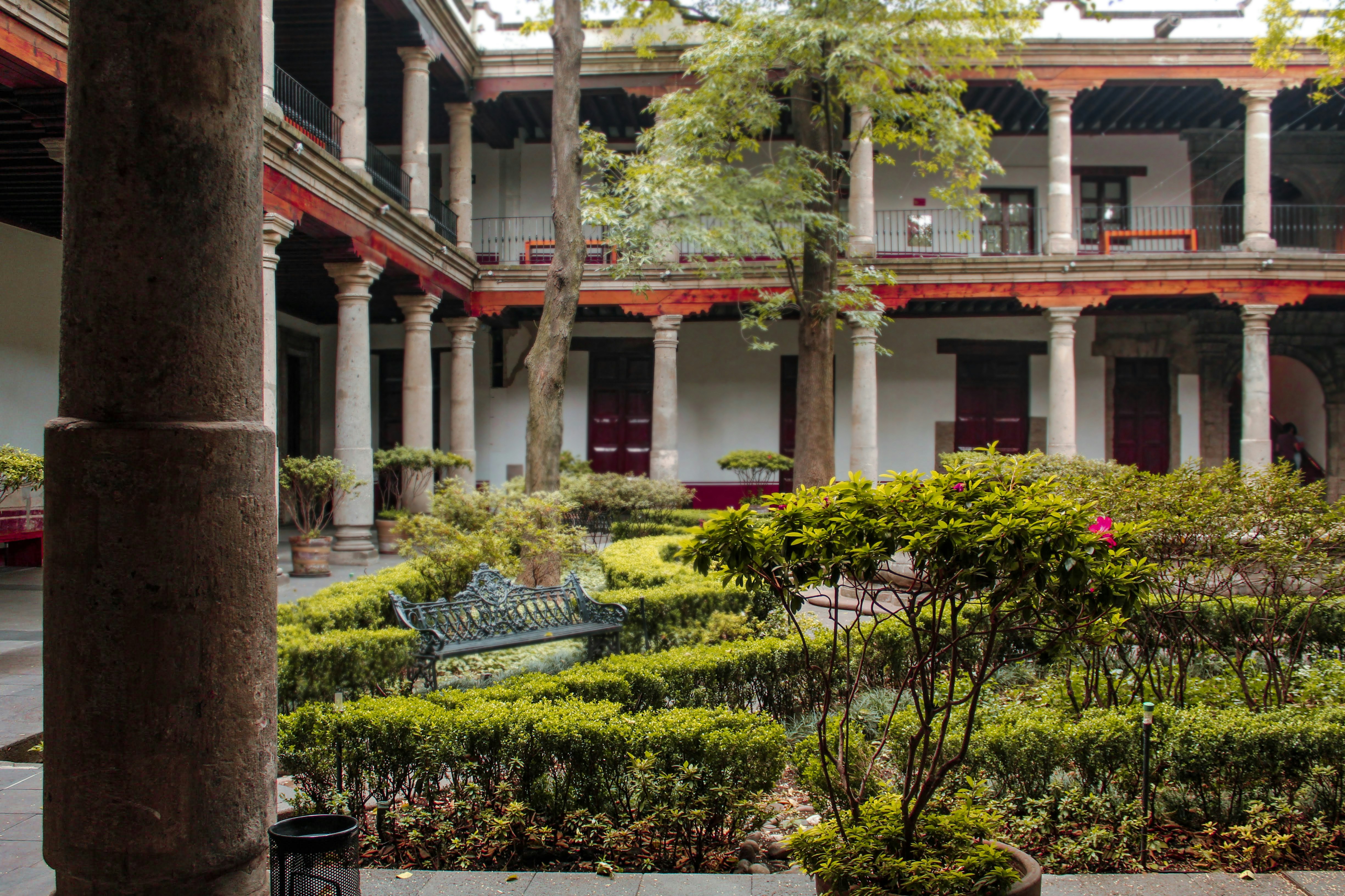 Lush courtyard garden surrounded by colonial architecture, featuring manicured hedges and a wrought-iron bench. A tranquil oasis invites reflection.