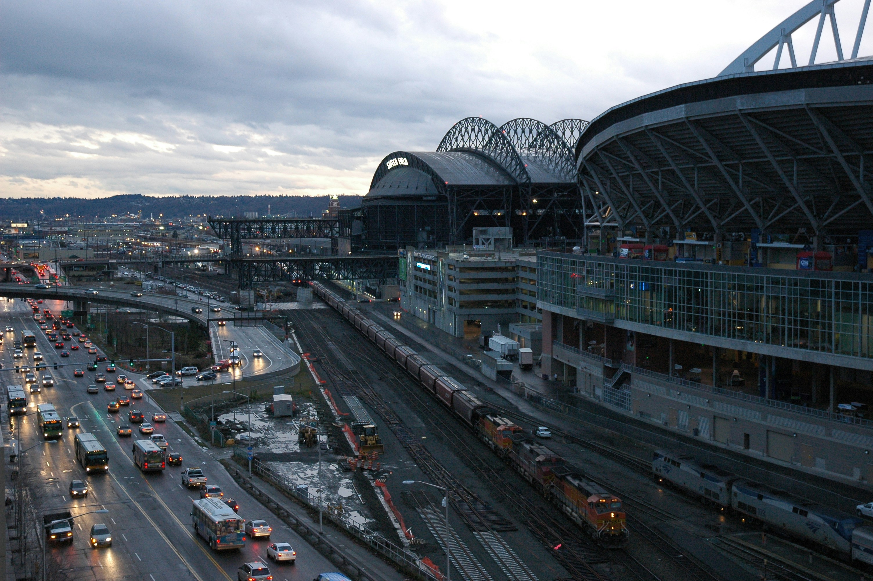Seattle's 3rd Avenue with busy traffic, sports arenas, and train tracks under an overcast winter evening sky.