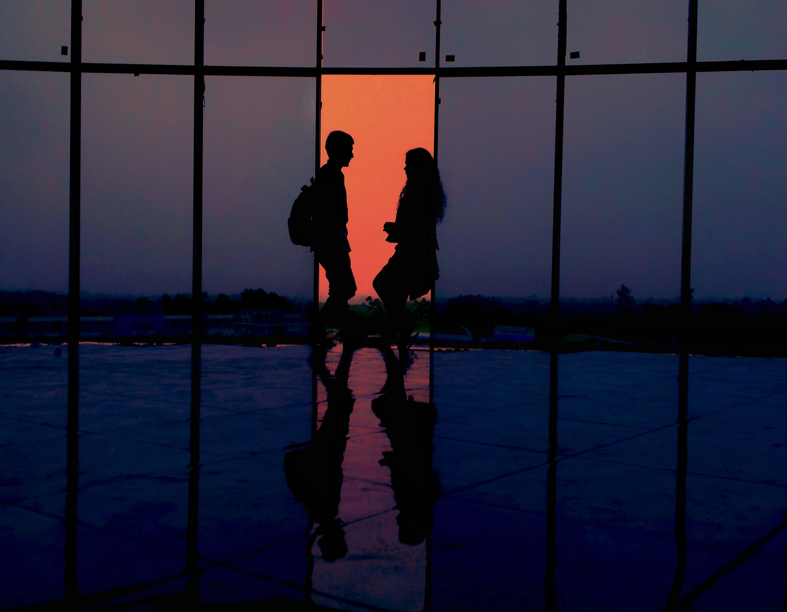 Two silhouetted figures converse against a vibrant sunset backdrop, reflected on a glossy floor.