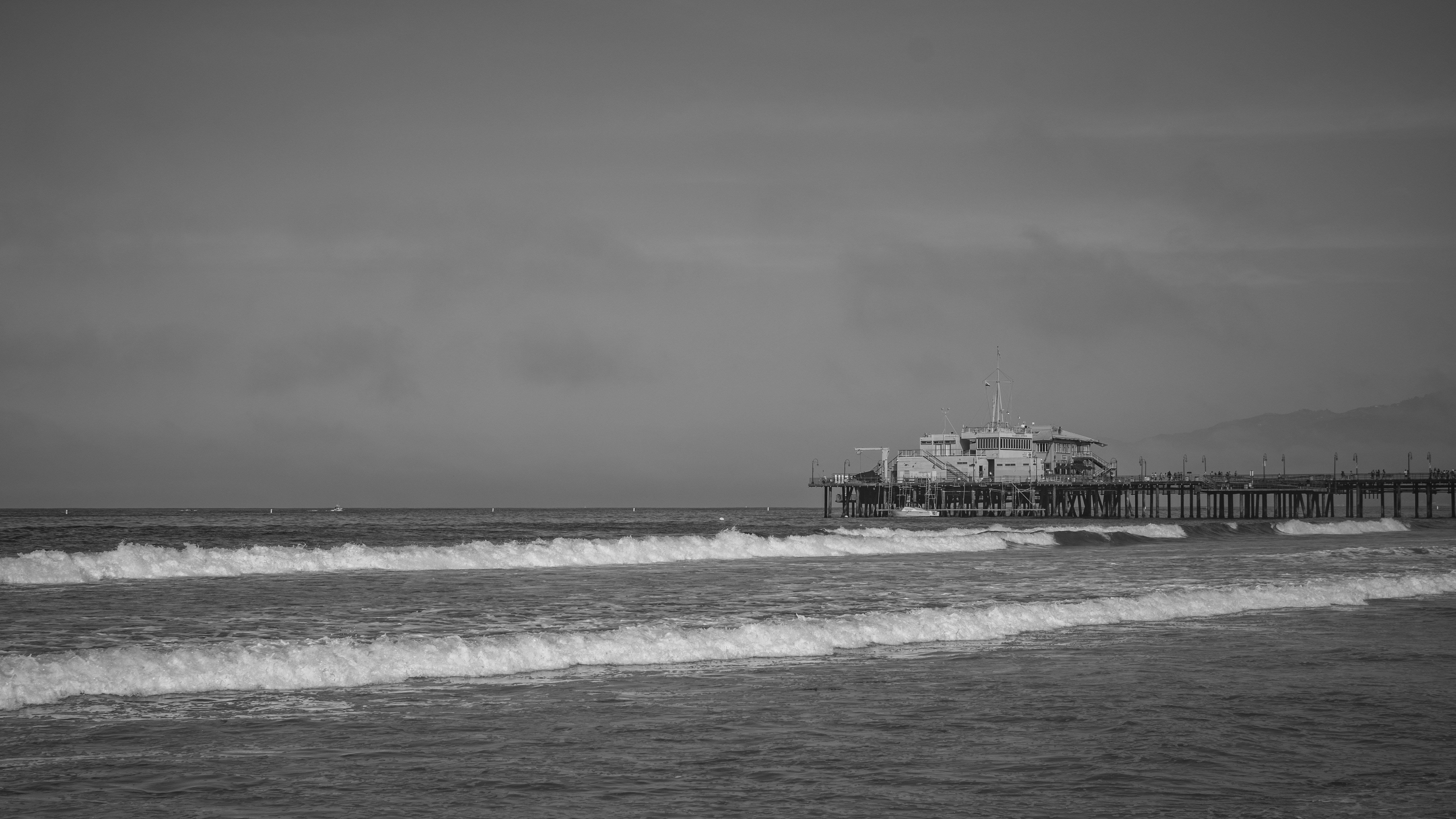 A weathered pier extends into the ocean under a cloudy sky, with gentle waves lapping at its base. The monochrome palette emphasizes the tranquil yet melancholic atmosphere.