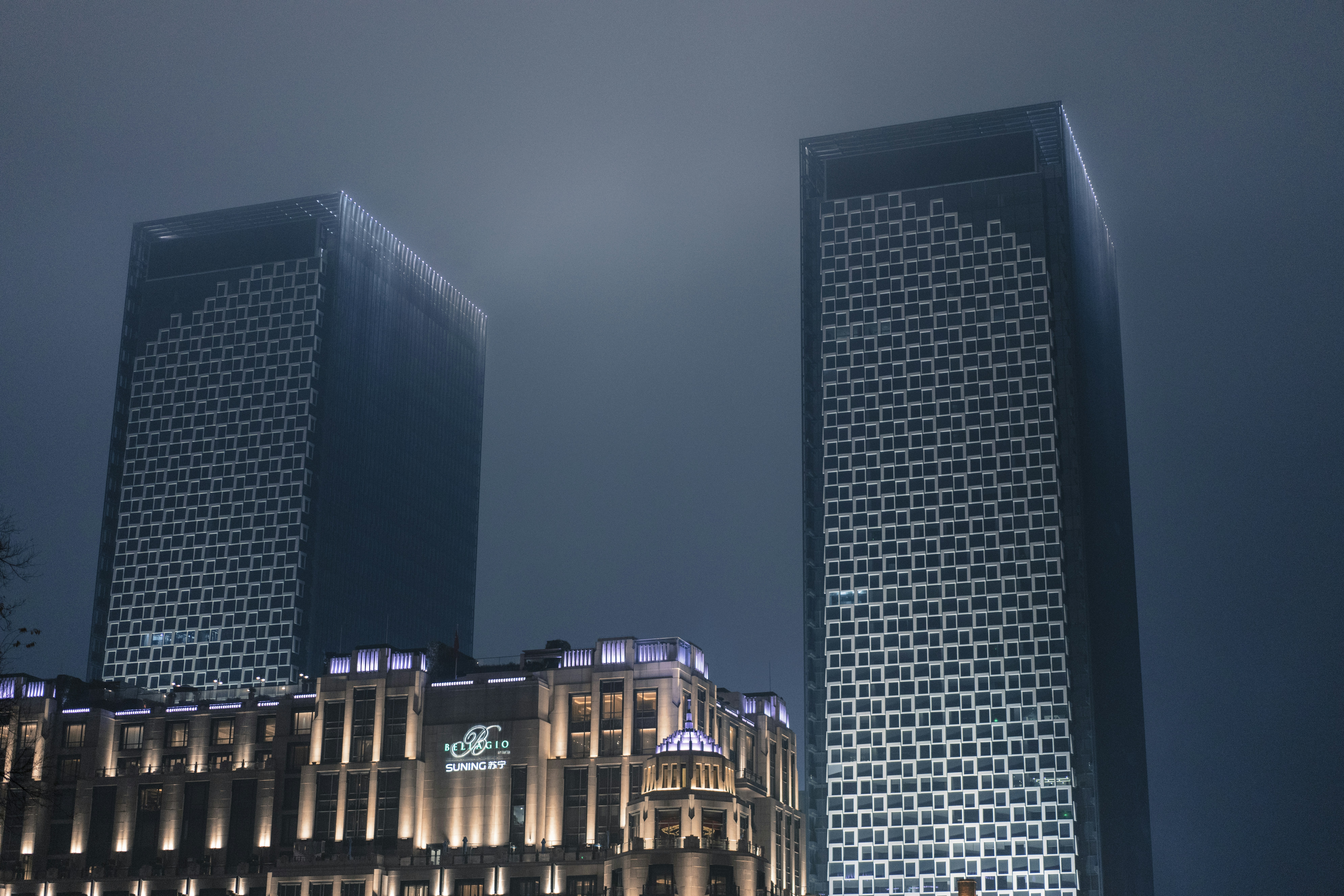 Two towering skyscrapers loom in a foggy urban landscape, illuminated by soft, ambient lighting. The contrasting architectural styles create a striking visual narrative.
