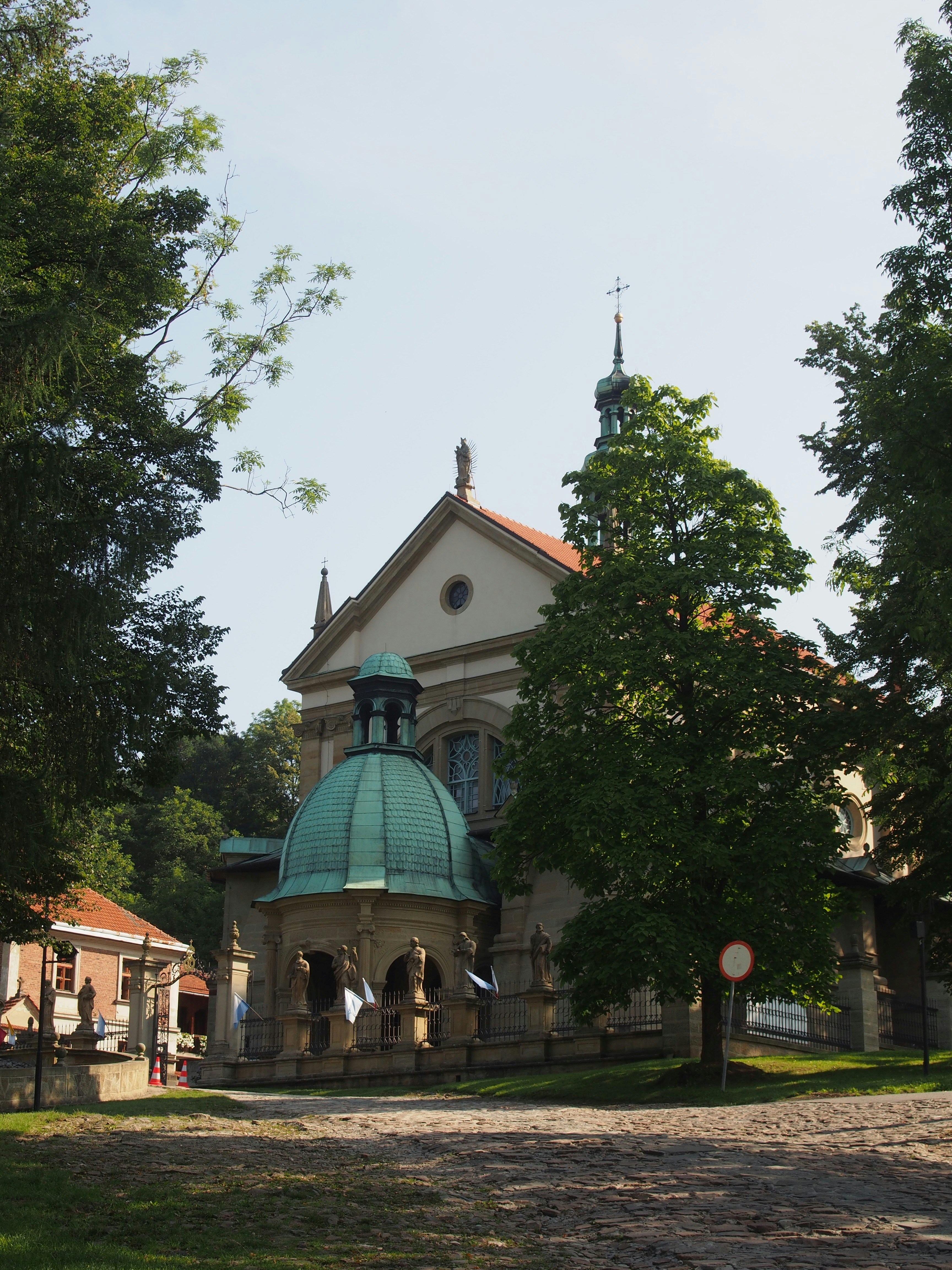 Sunlit view of a historic church with a verdigris copper dome, stone facade, and surrounding trees; cobblestone foreground leads to the entrance.