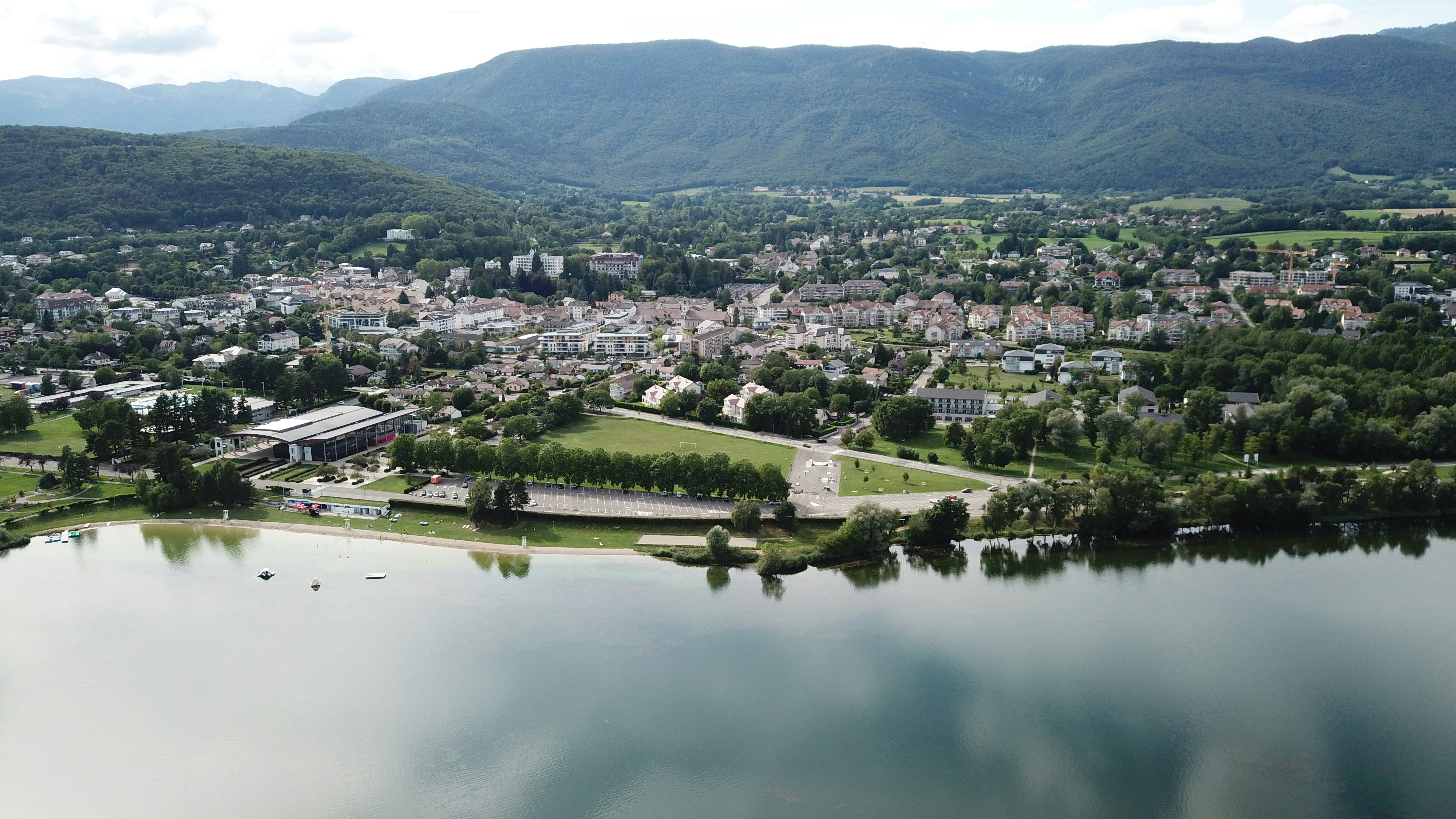 Aerial view of a serene village nestled beside a lake with lush greenery and distant mountains under a cloudy sky.