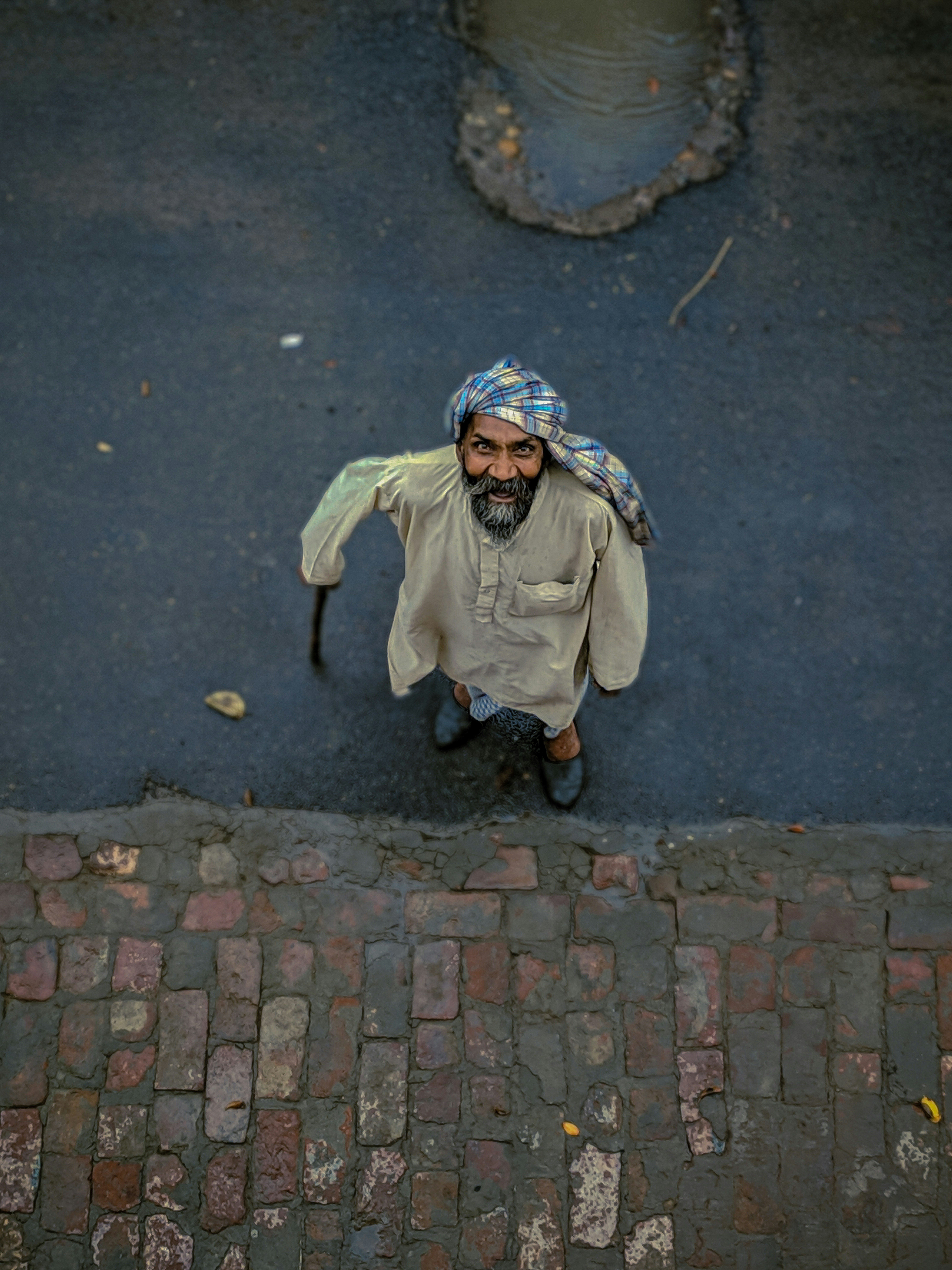 Elderly man with a cane stands on a cobbled street, gazing upward with a thoughtful expression. His traditional attire and weathered features tell a story of experience.
