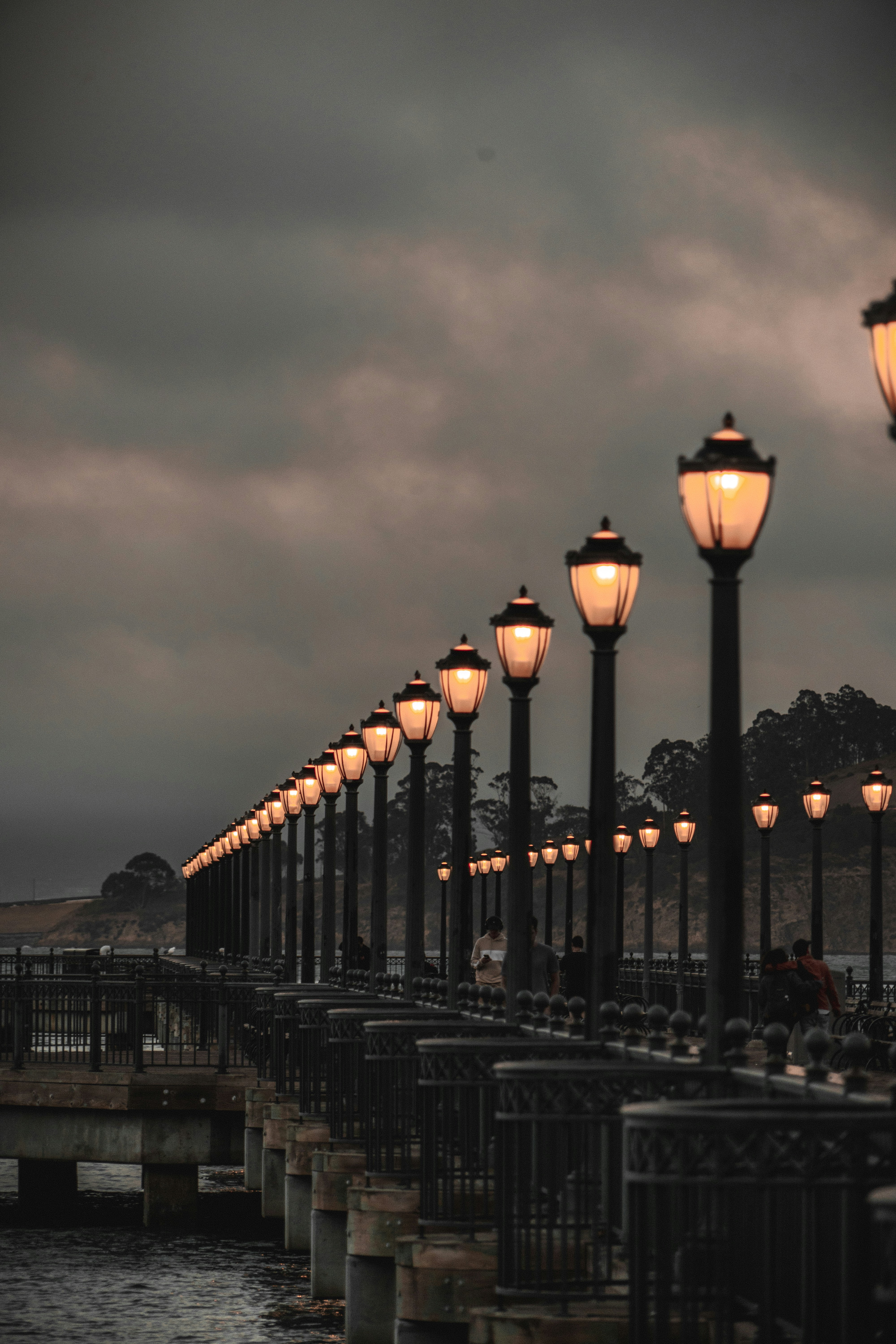A row of glowing lampposts lines a pier, casting warm light against a backdrop of dark, cloudy skies. The scene evokes a sense of calm amidst the impending storm.