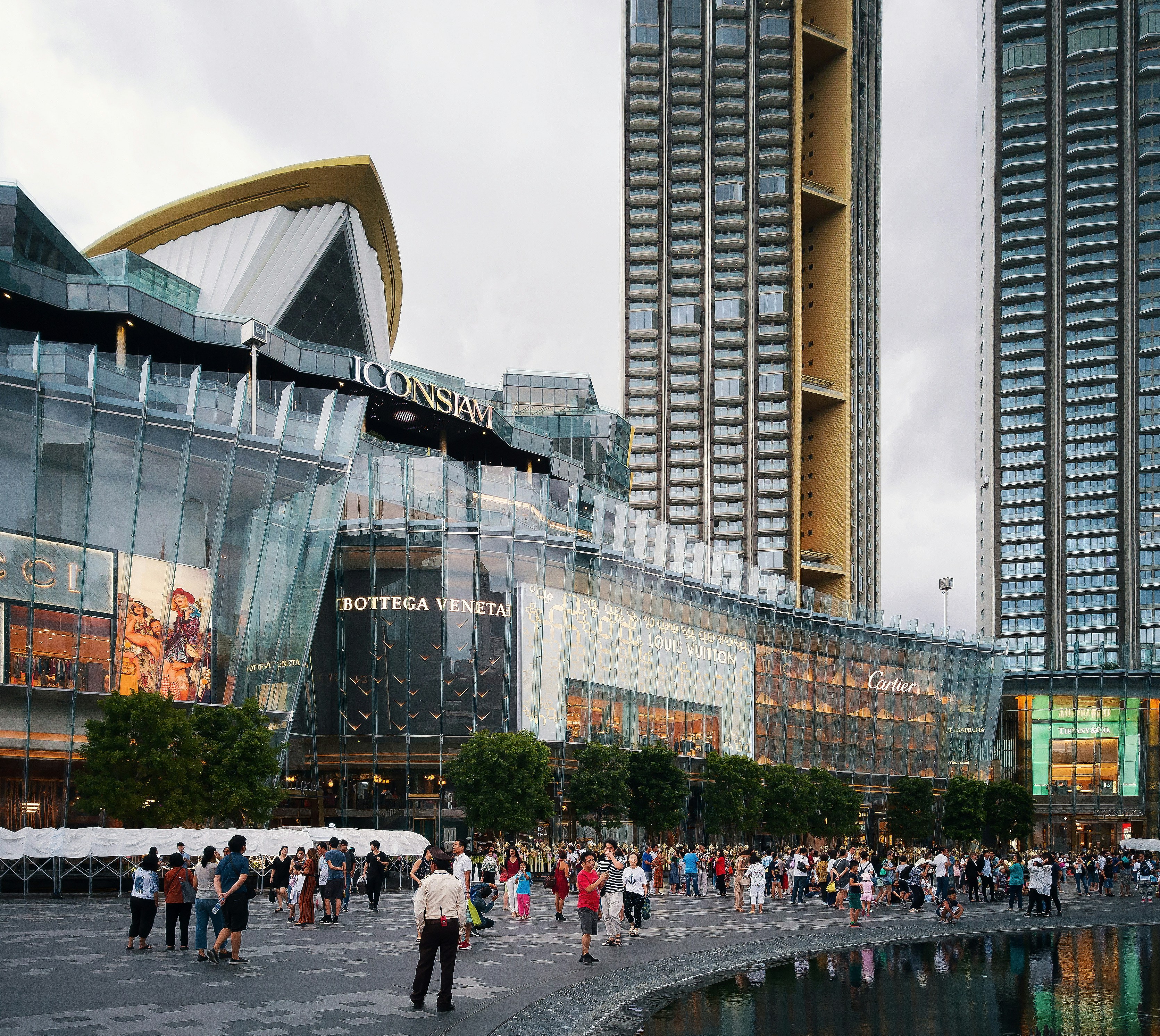 Crowds gather near a modern shopping mall with towering skyscrapers in the background under a cloudy sky.