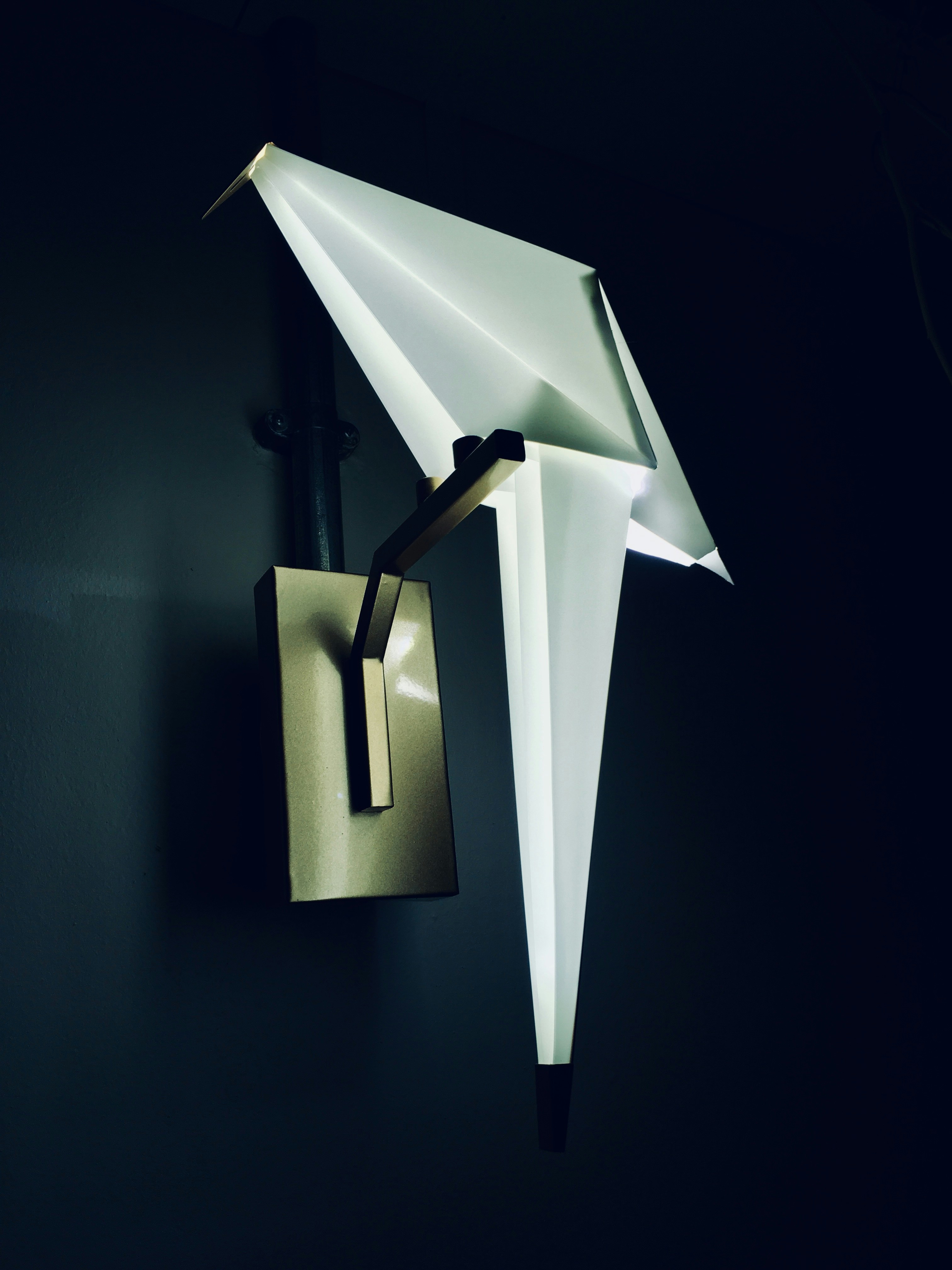 Angular wall lamp with a geometric, origami-inspired shade glows against a dark blue wall in a moody indoor photograph.