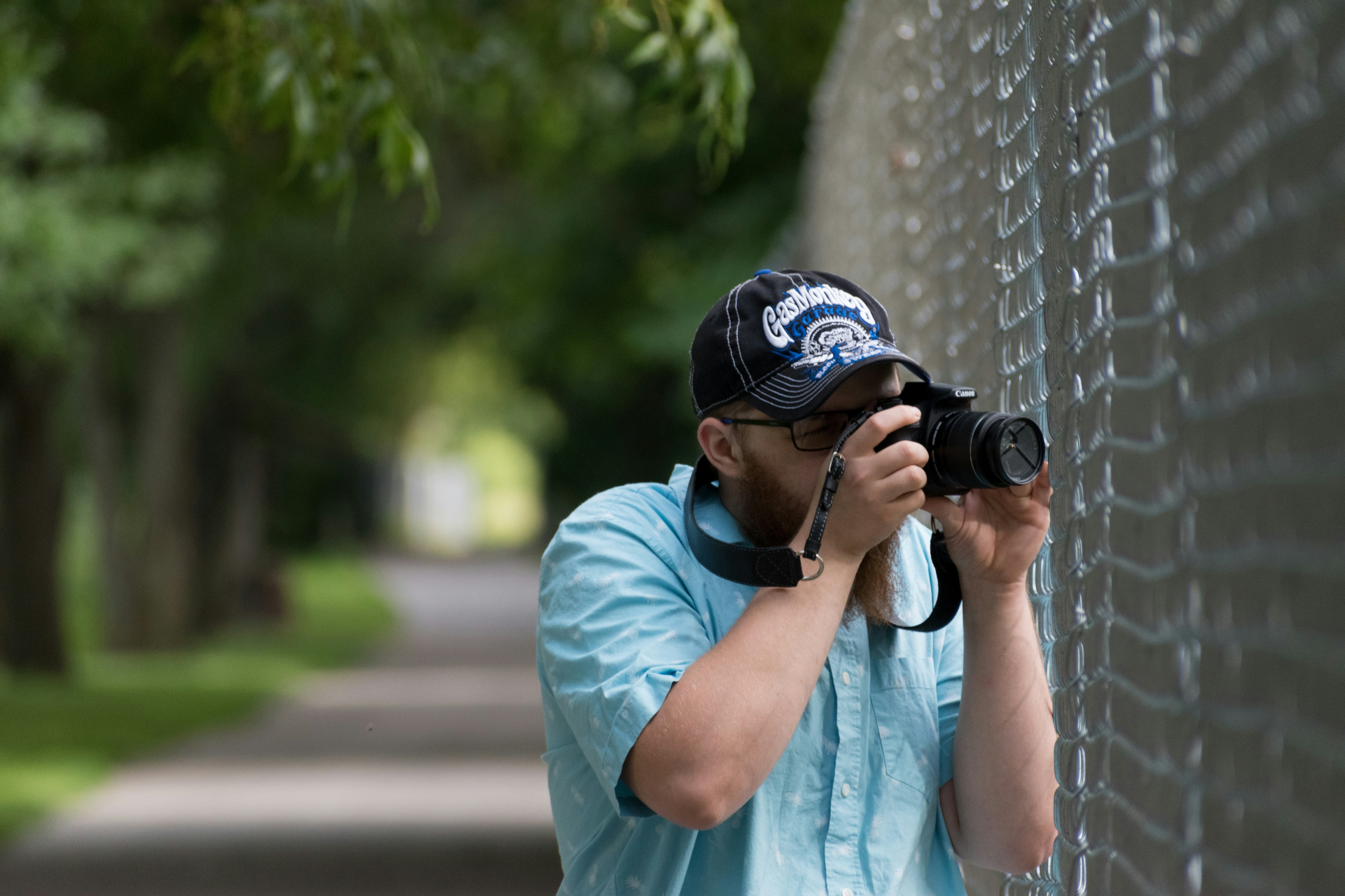 A photographer in a light blue shirt captures an image while leaning against a chain-link fence, surrounded by a lush green backdrop. The scene conveys a sense of concentration and creativity.