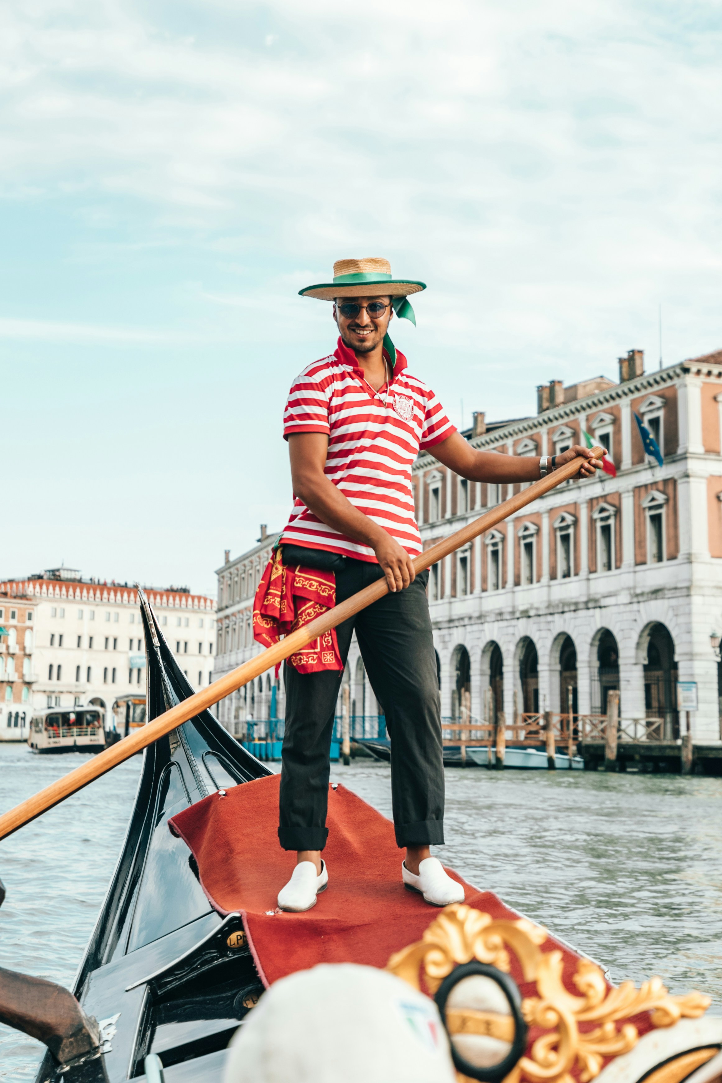 Gondolier in a striped shirt and hat skillfully maneuvers a gondola through the canals of Venice, with historic buildings lining the waterfront.