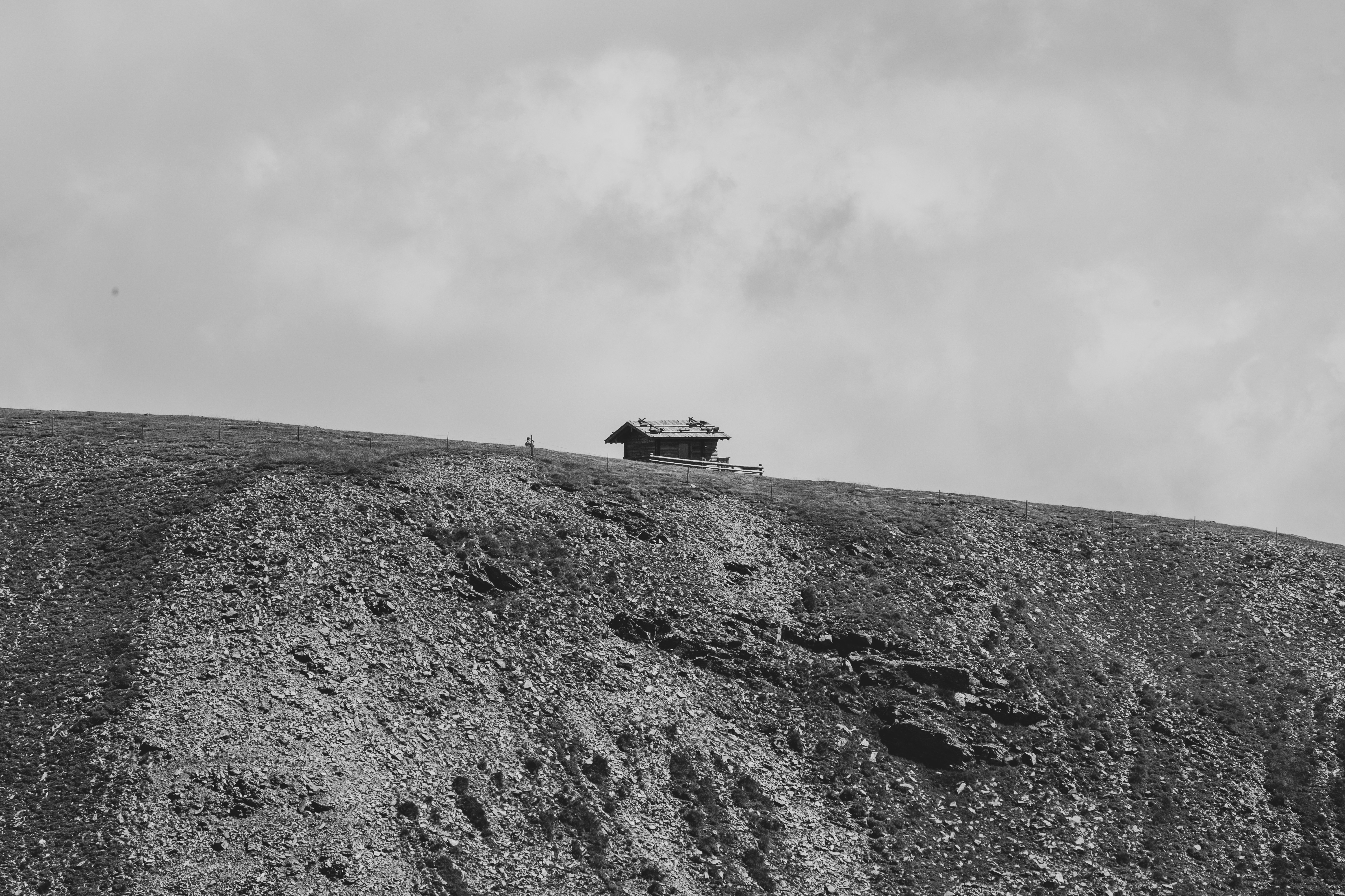 A lone cabin perched atop a rugged hill, surrounded by a barren landscape under a cloudy sky.