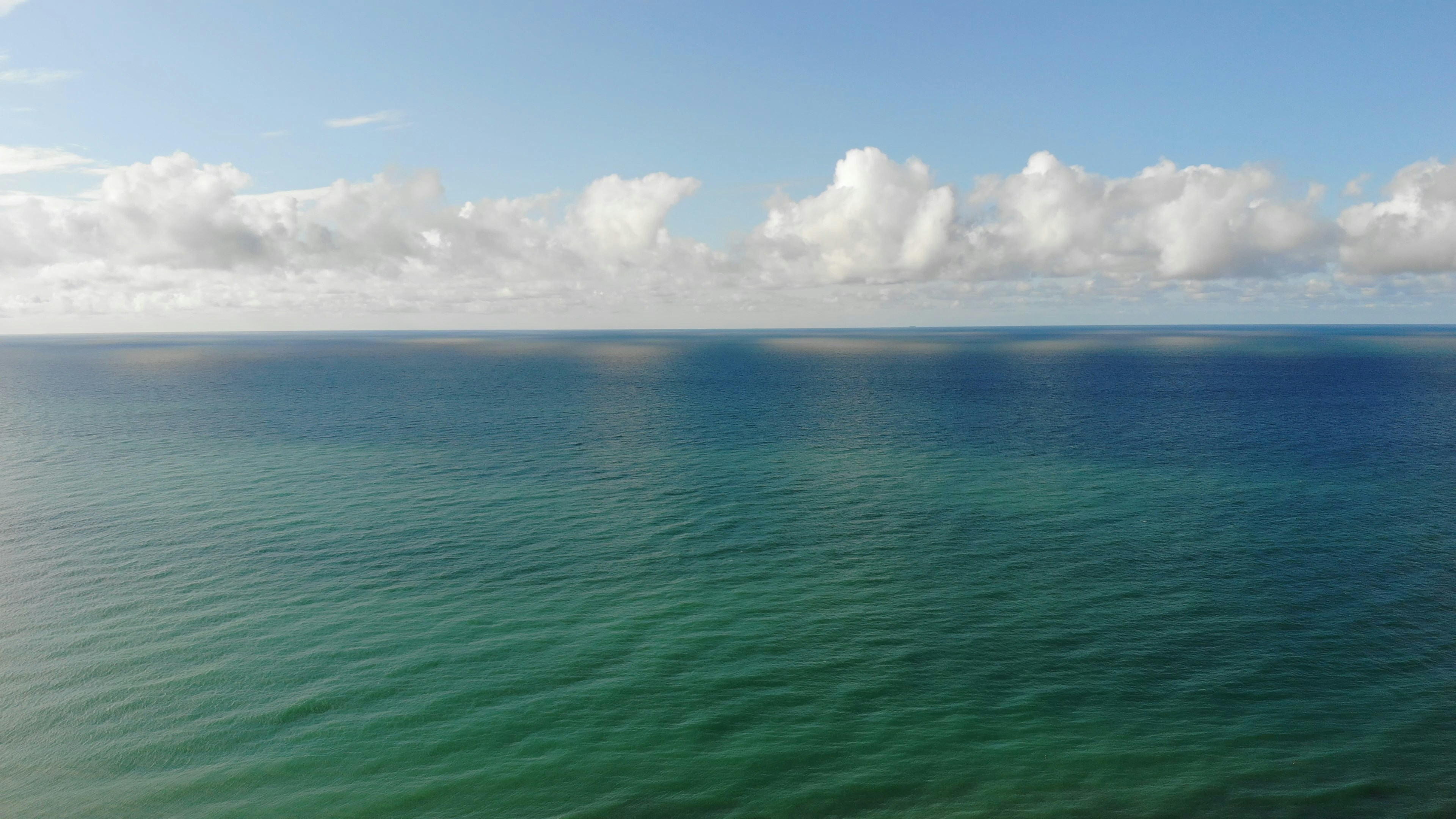 Vast ocean expanse meets a horizon dotted with fluffy clouds, showcasing a gradient of blues and greens. The tranquil waters reflect the sky's hues beautifully.