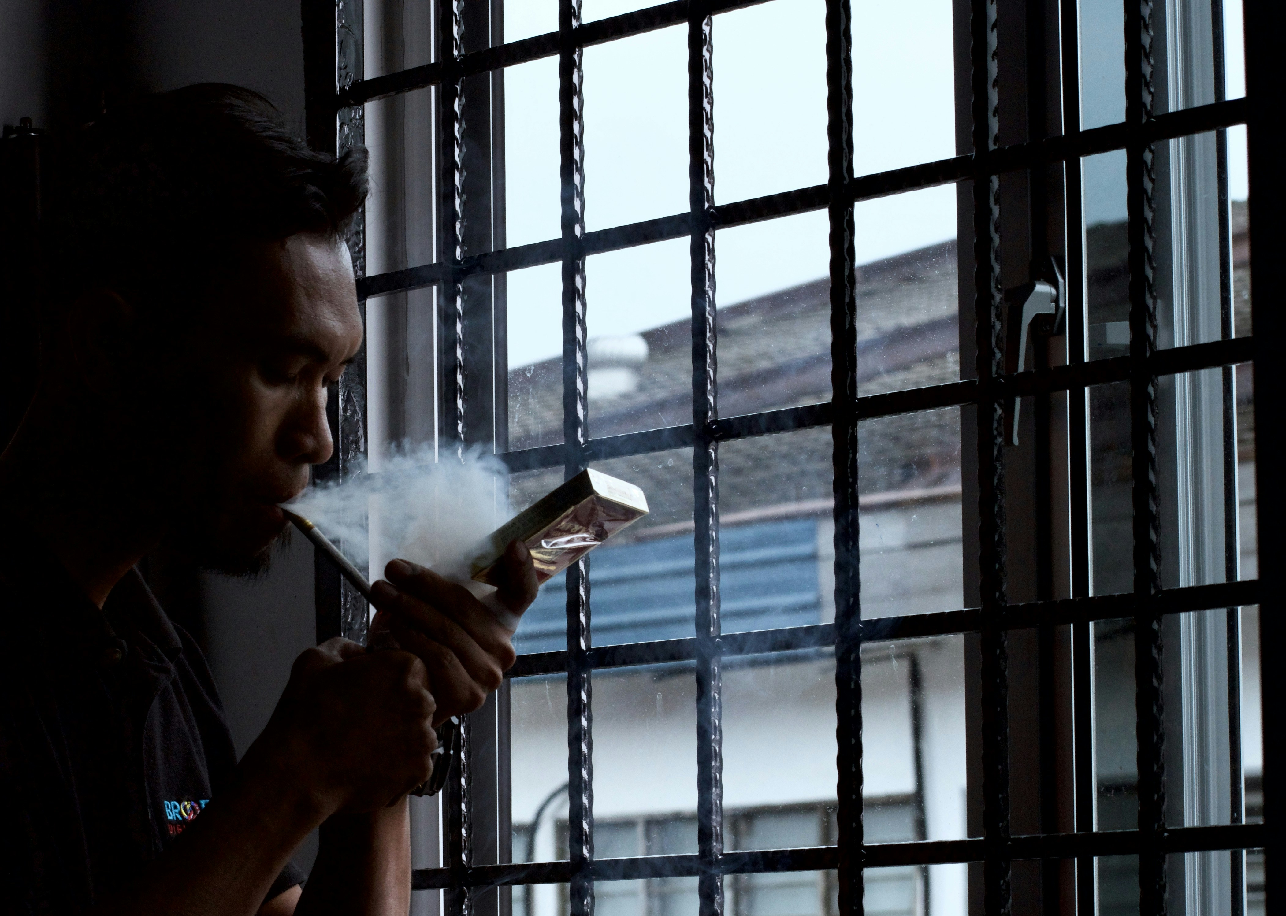 Man exhaling vapor while leaning against a window with a grid pattern, creating a contemplative atmosphere.