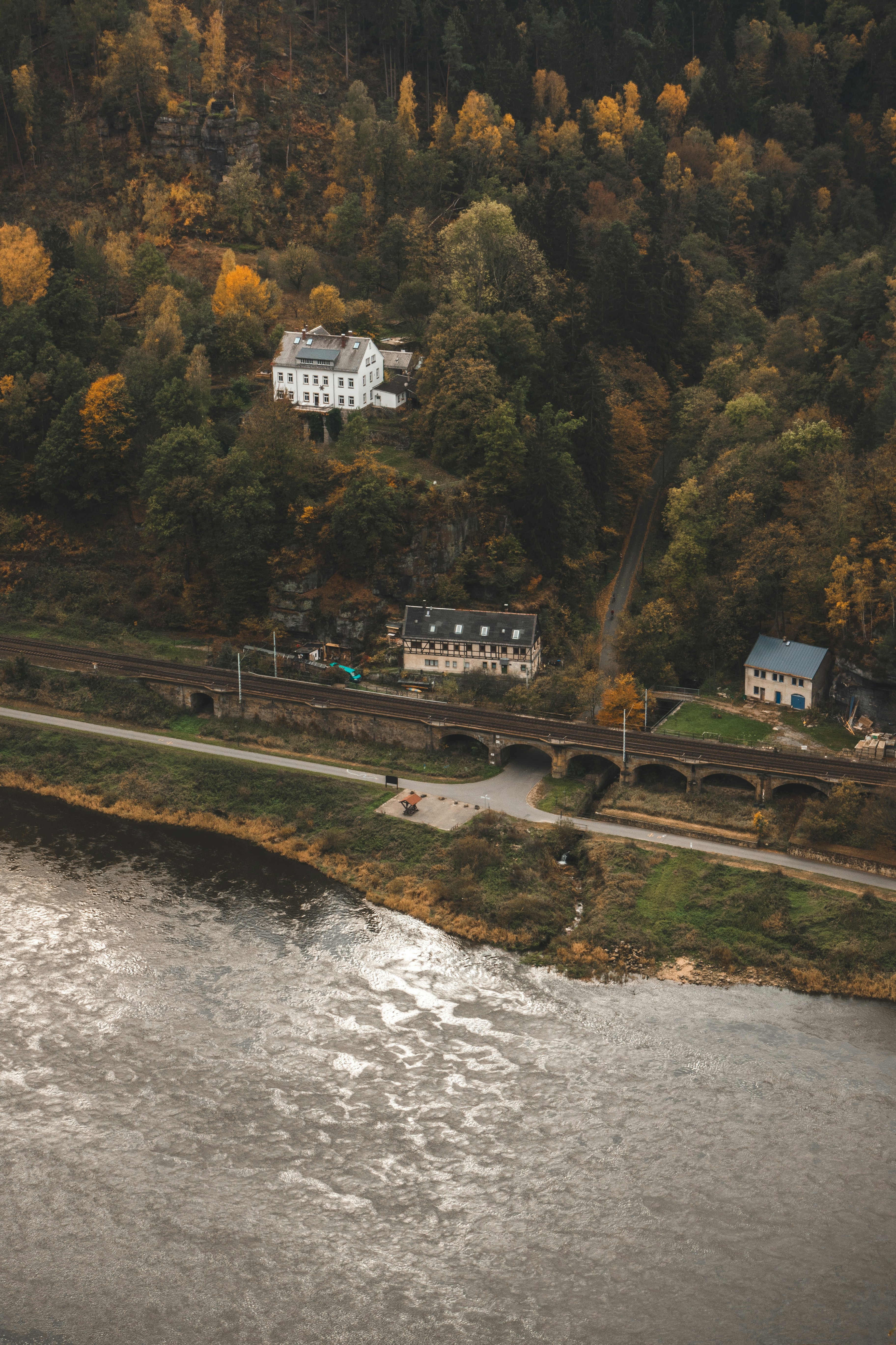 Aerial view of a serene riverside scene featuring a cluster of houses surrounded by autumn foliage, with a winding road and a bridge crossing over the river.