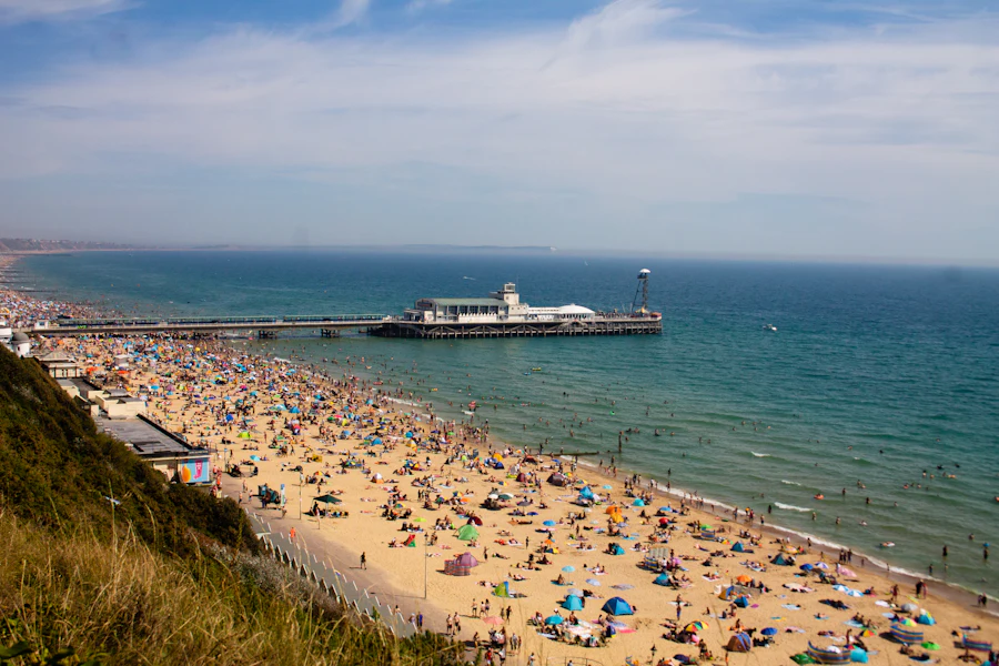 Crowded sandy beach beside a calm blue sea, with a long pier extending into the water under a hazy sky. Distant shoreline and grassy cliff edge frame the scene.