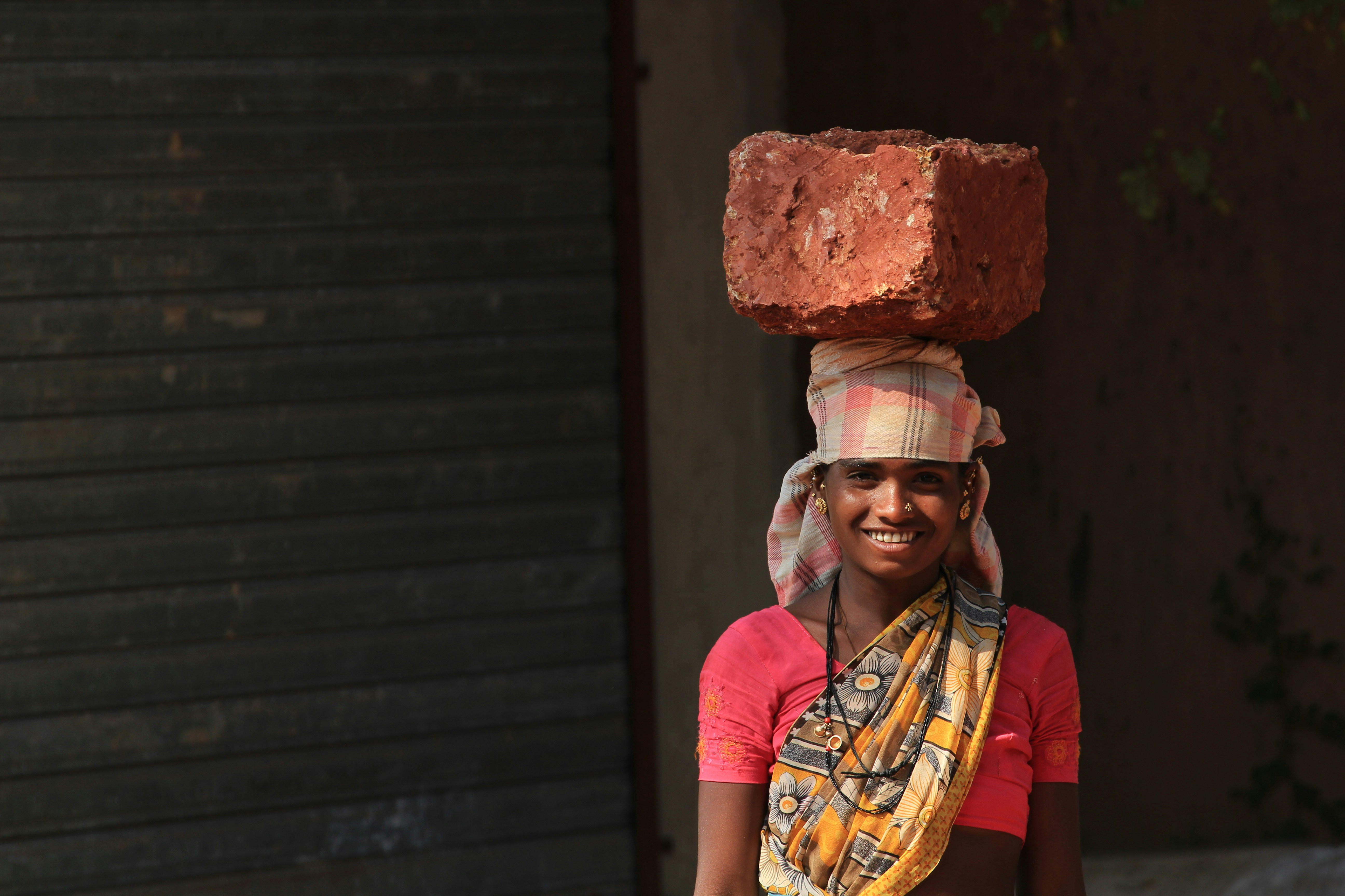 A young individual proudly balancing a brick on their head, showcasing determination amidst a rustic backdrop. The vibrant attire contrasts with the subdued setting.