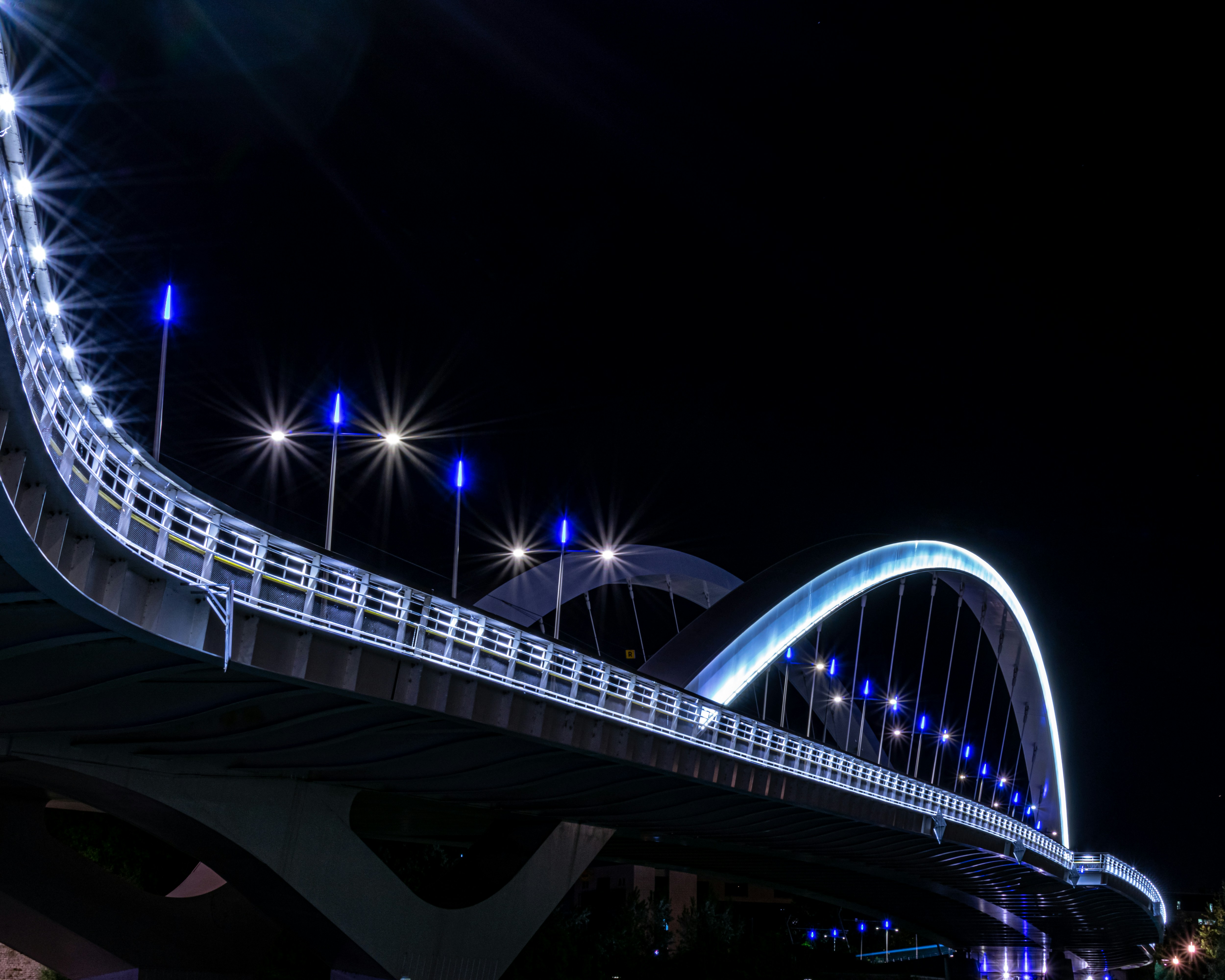 A modern bridge illuminated by vibrant blue and white lights against a dark sky, showcasing its architectural design and nighttime ambiance.
