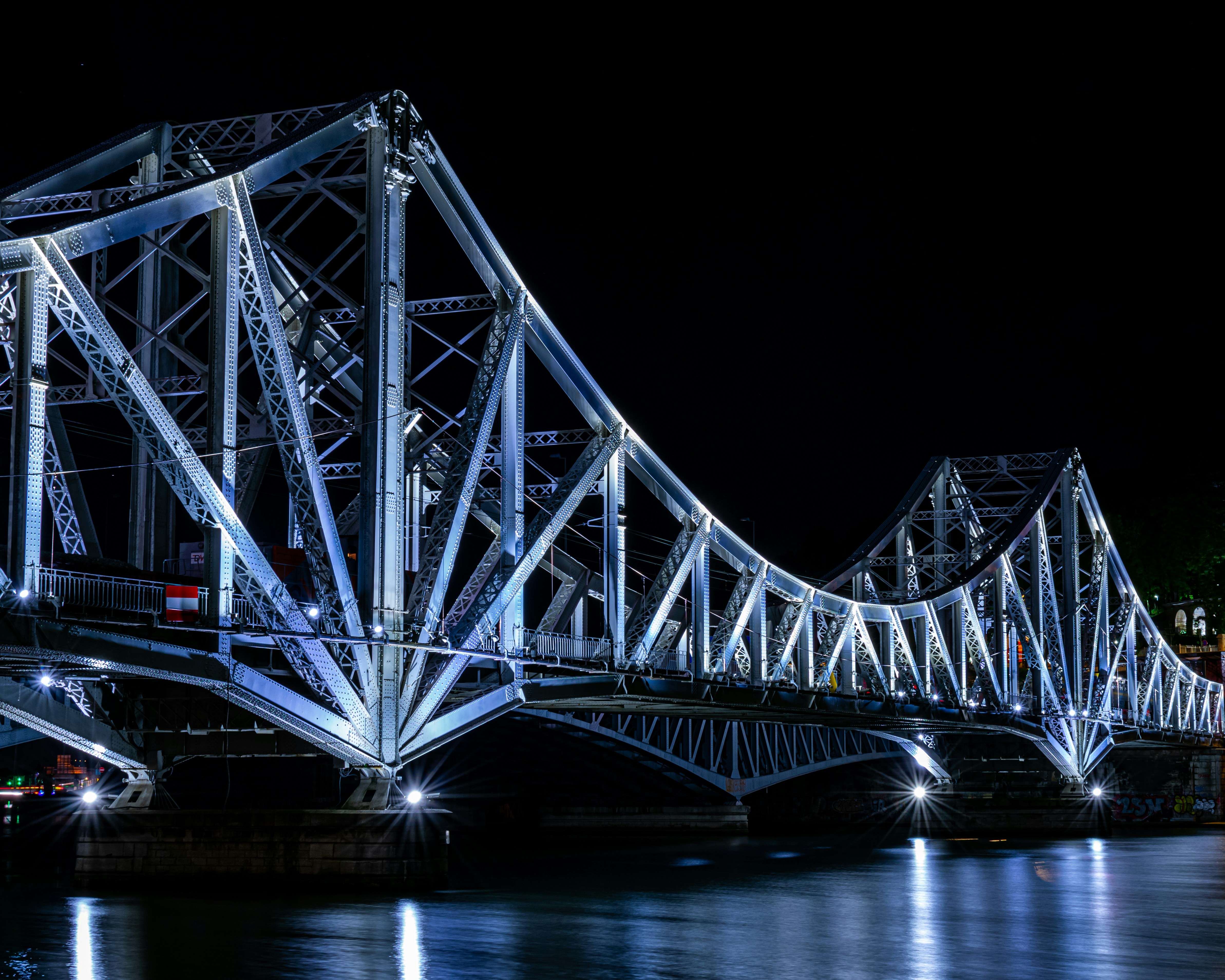 A striking bridge illuminated against the night sky, showcasing intricate steelwork and reflections in the water below.