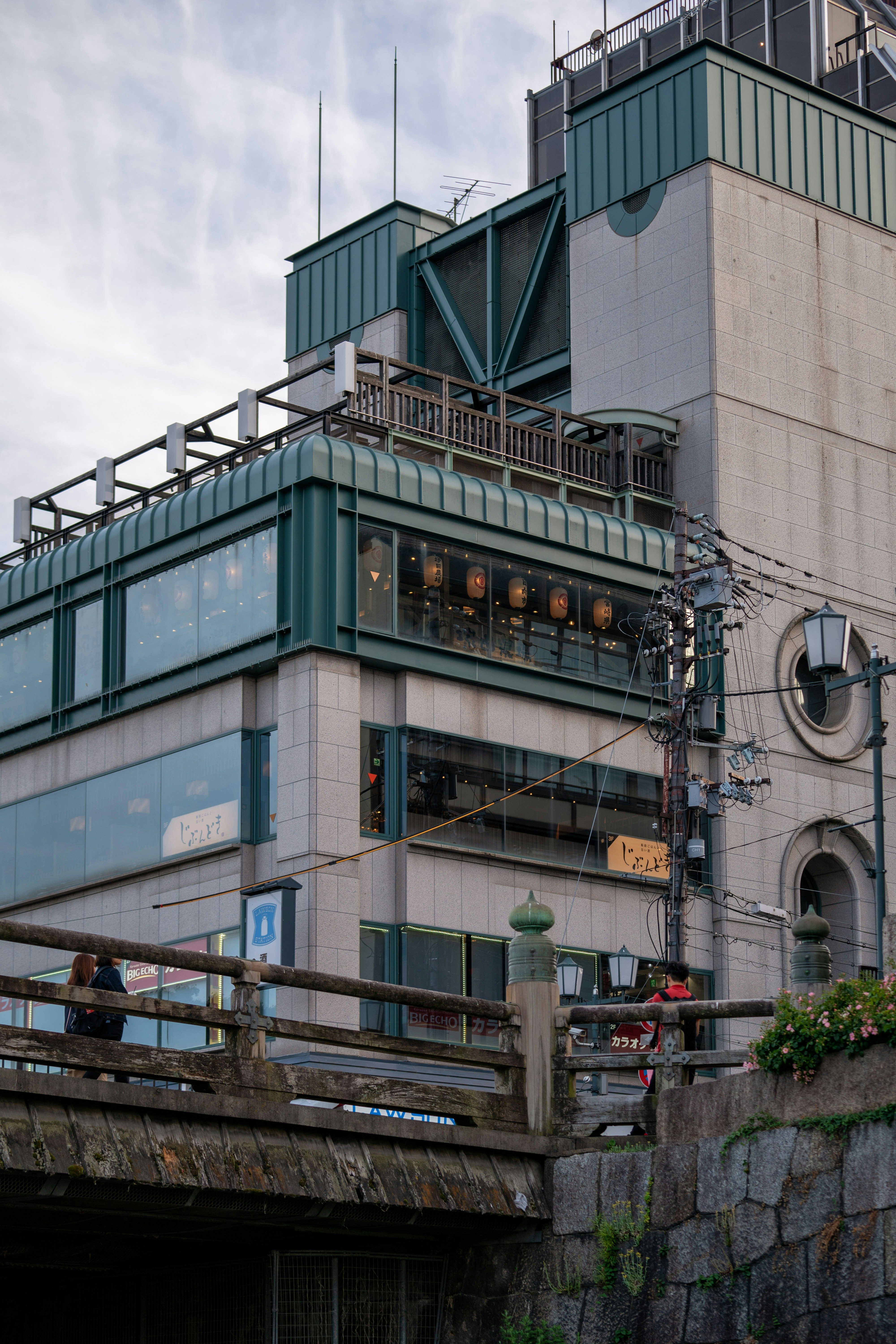 Contemporary building with glass facades and decorative elements, juxtaposed against a rustic stone bridge and street lamps.