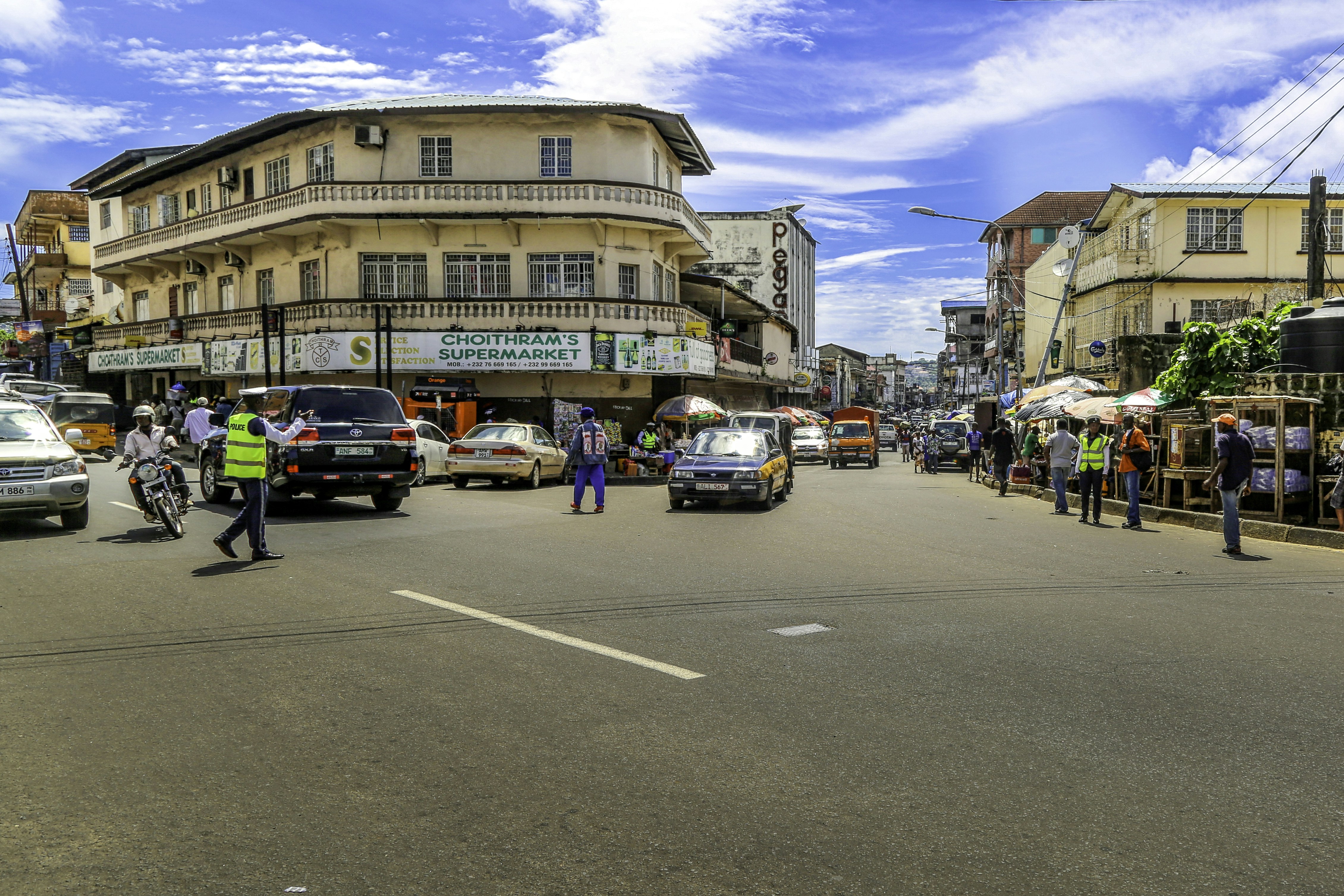 Busy street scene showcasing diverse vehicles and pedestrians at a bustling intersection, with a supermarket prominently featured. 