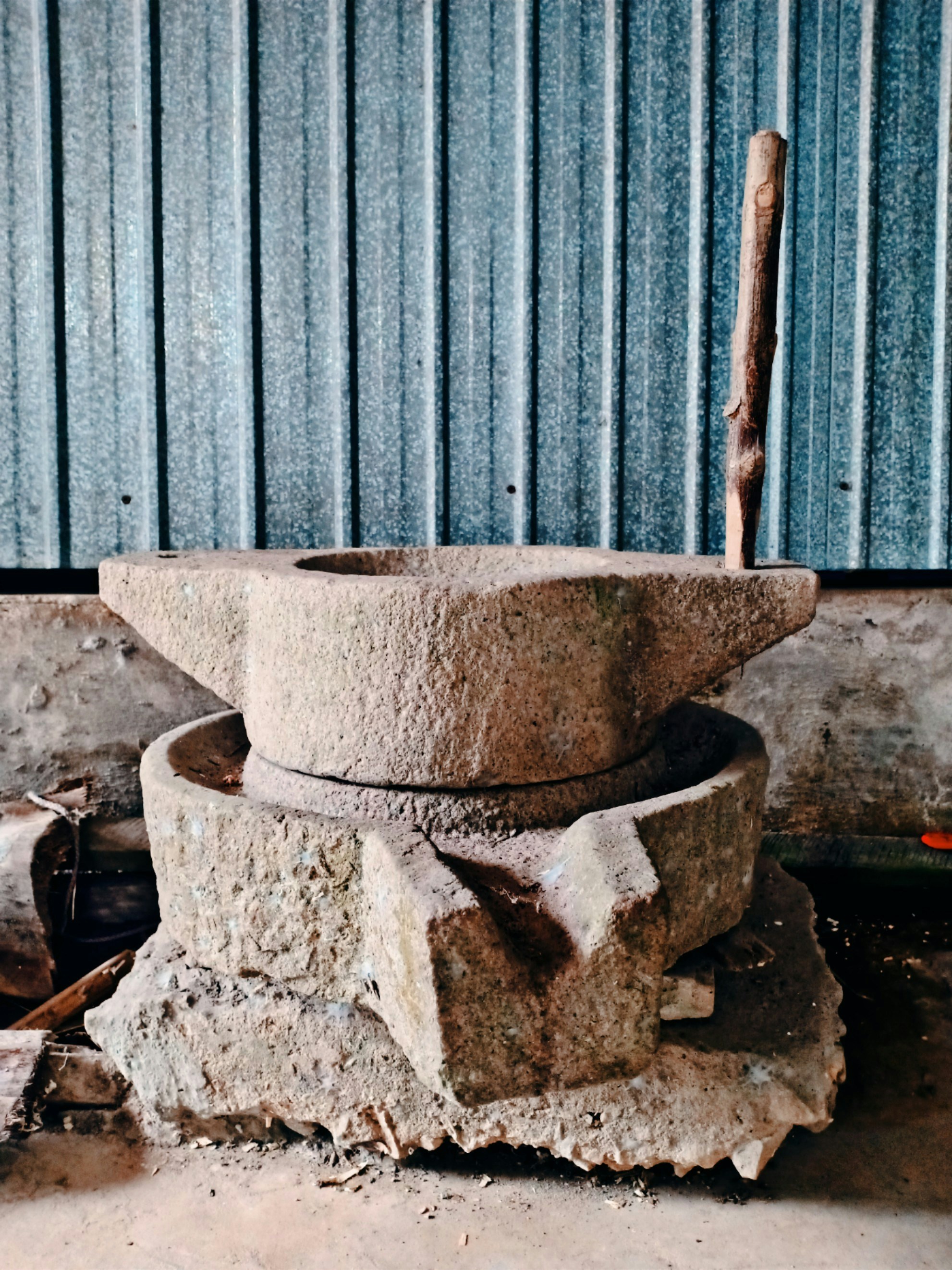 A rustic stone mortar and pestle sit atop a weathered base against a corrugated metal wall; muted earthy tones convey a worn, workshop look.