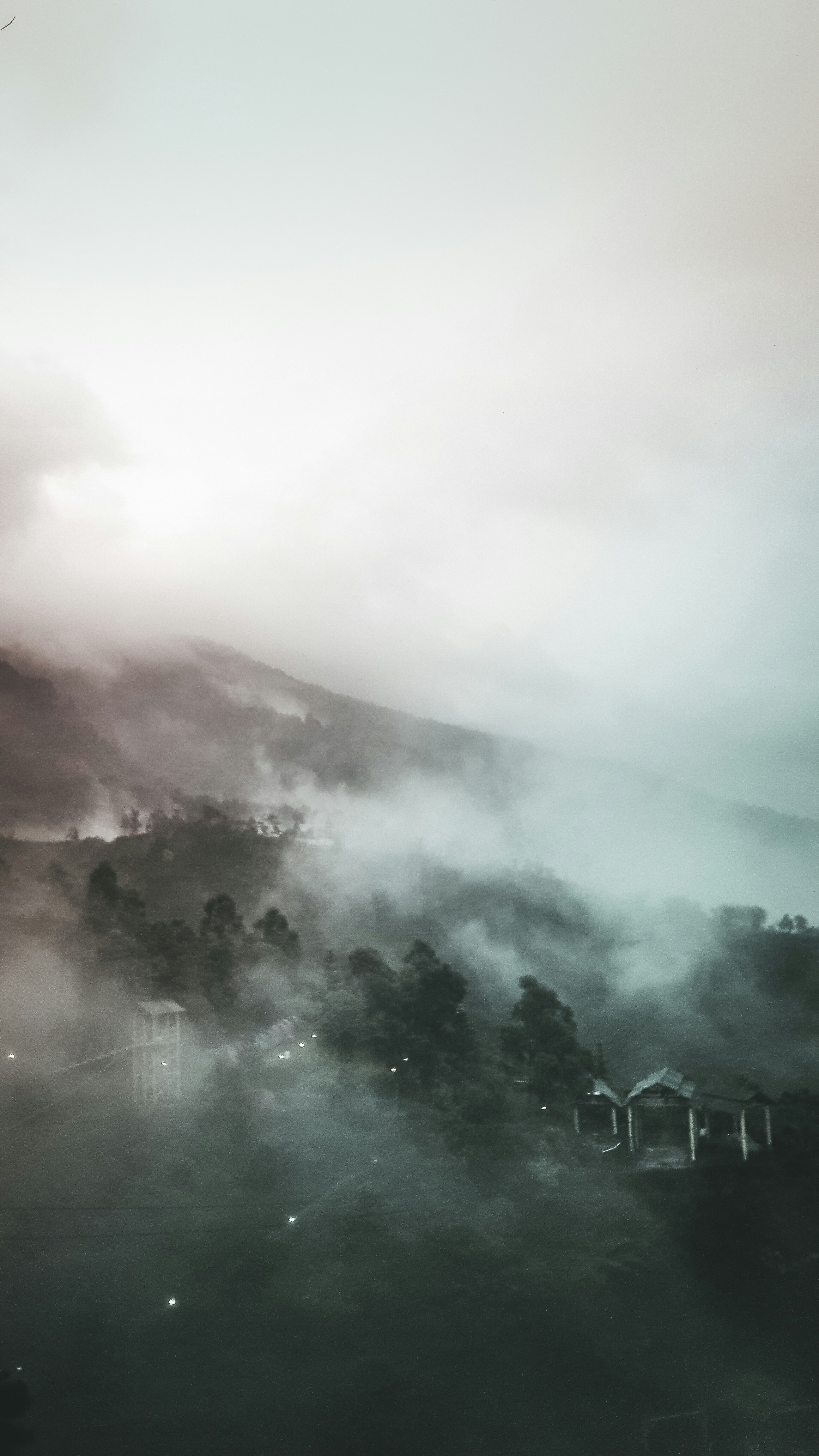 Fog envelops a mountainside with scattered trees and structures beneath a pale sky.