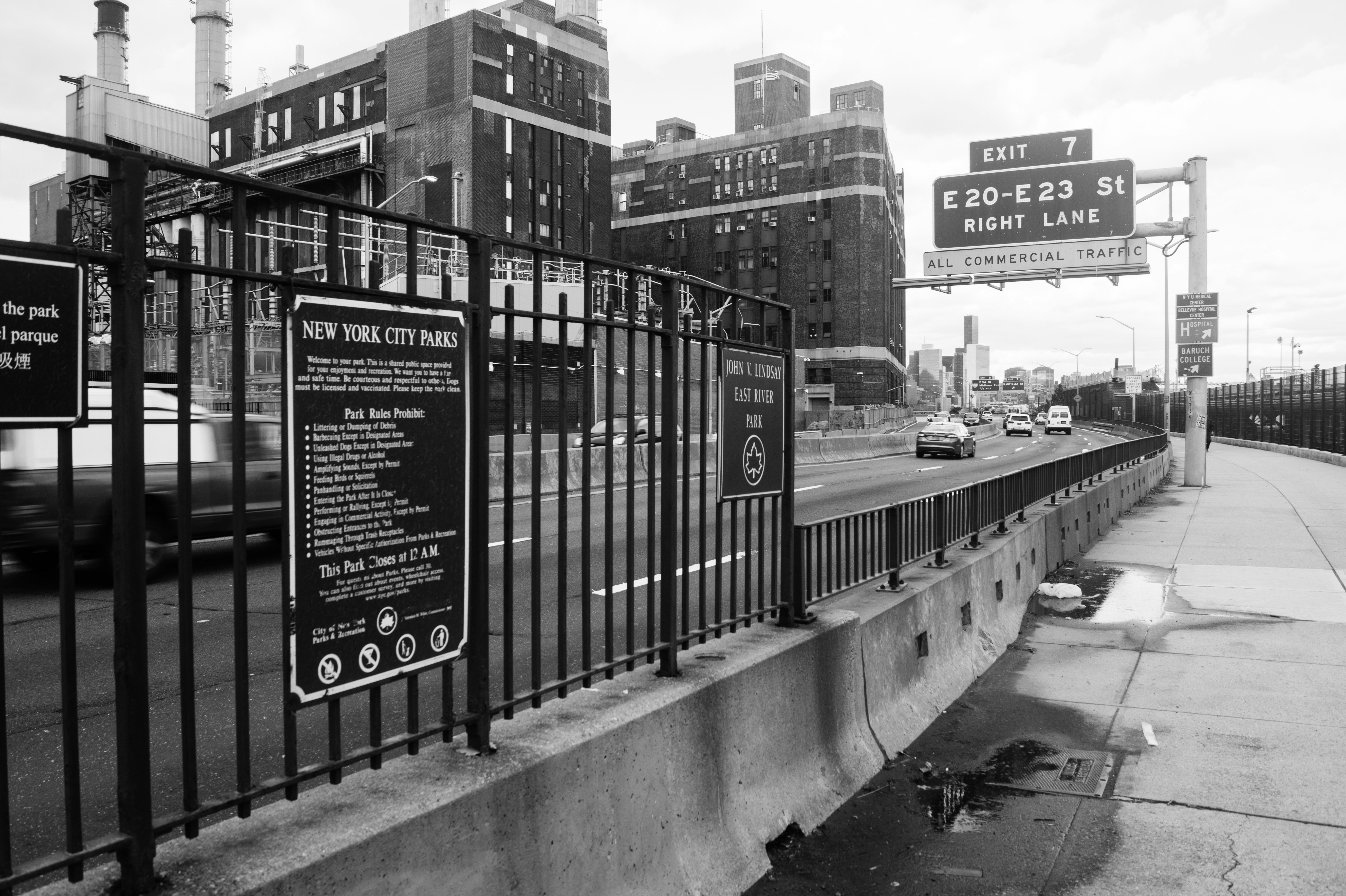 Black and white photograph showcasing a city street with a park sign and traffic signs, emphasizing urban infrastructure.