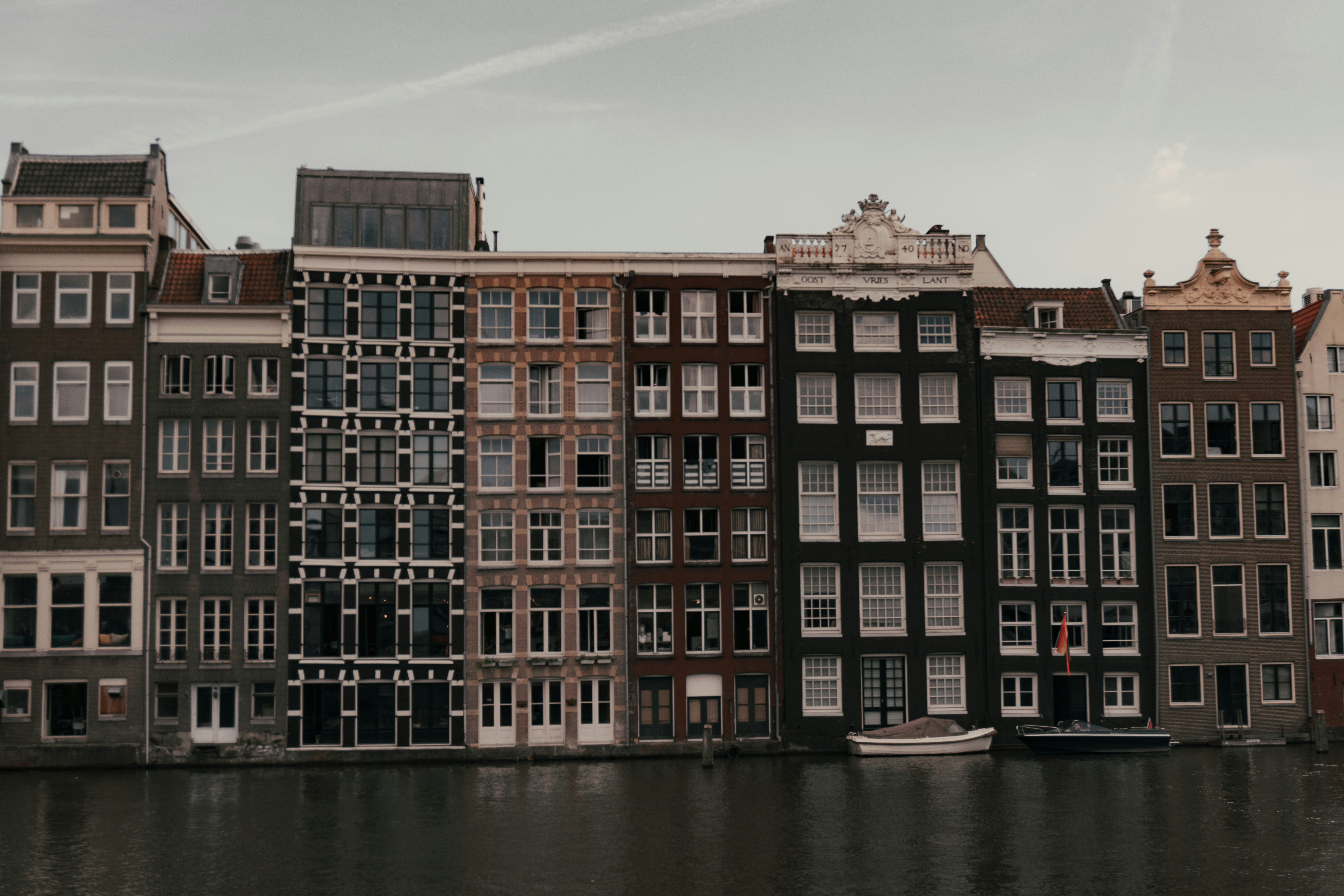 Historic Amsterdam buildings reflect in the tranquil canal water under a cloudy sky.