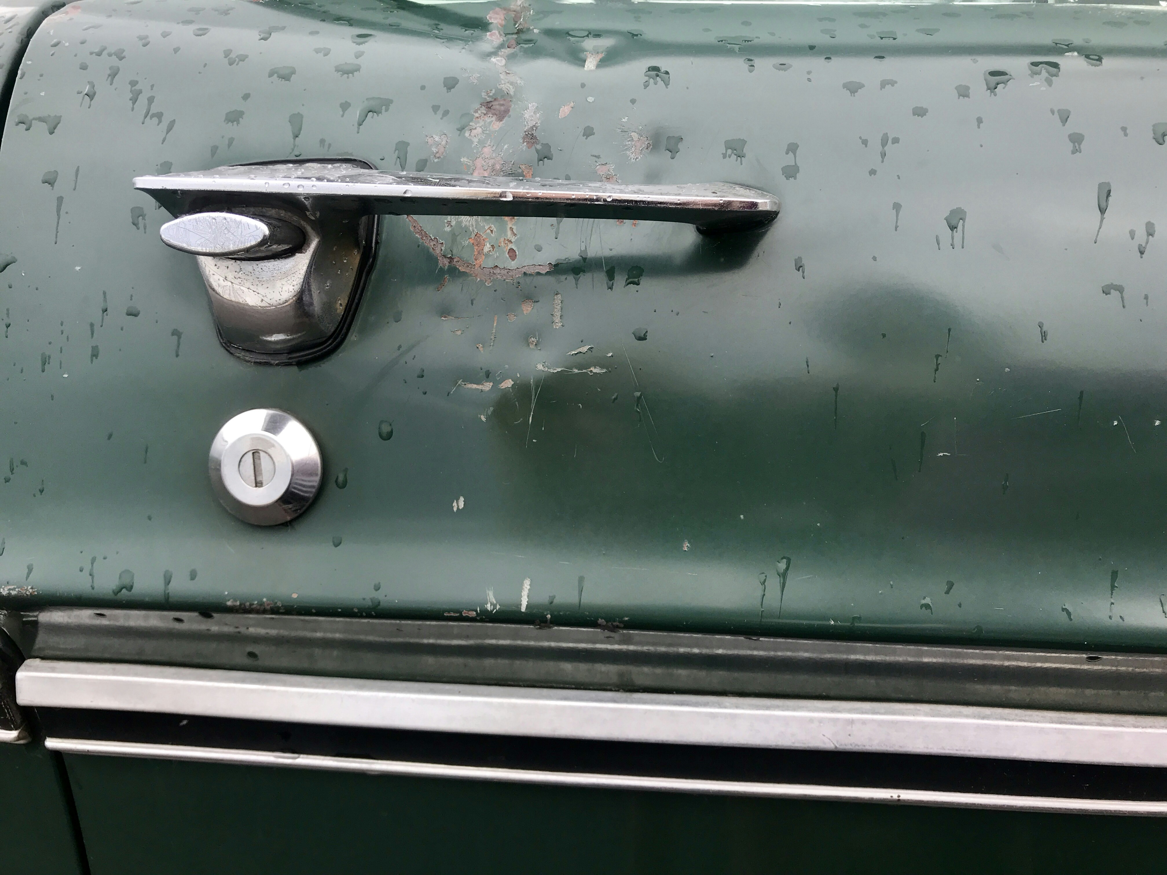 Close-up of a classic car's green door with chrome handle and visible rust marks.