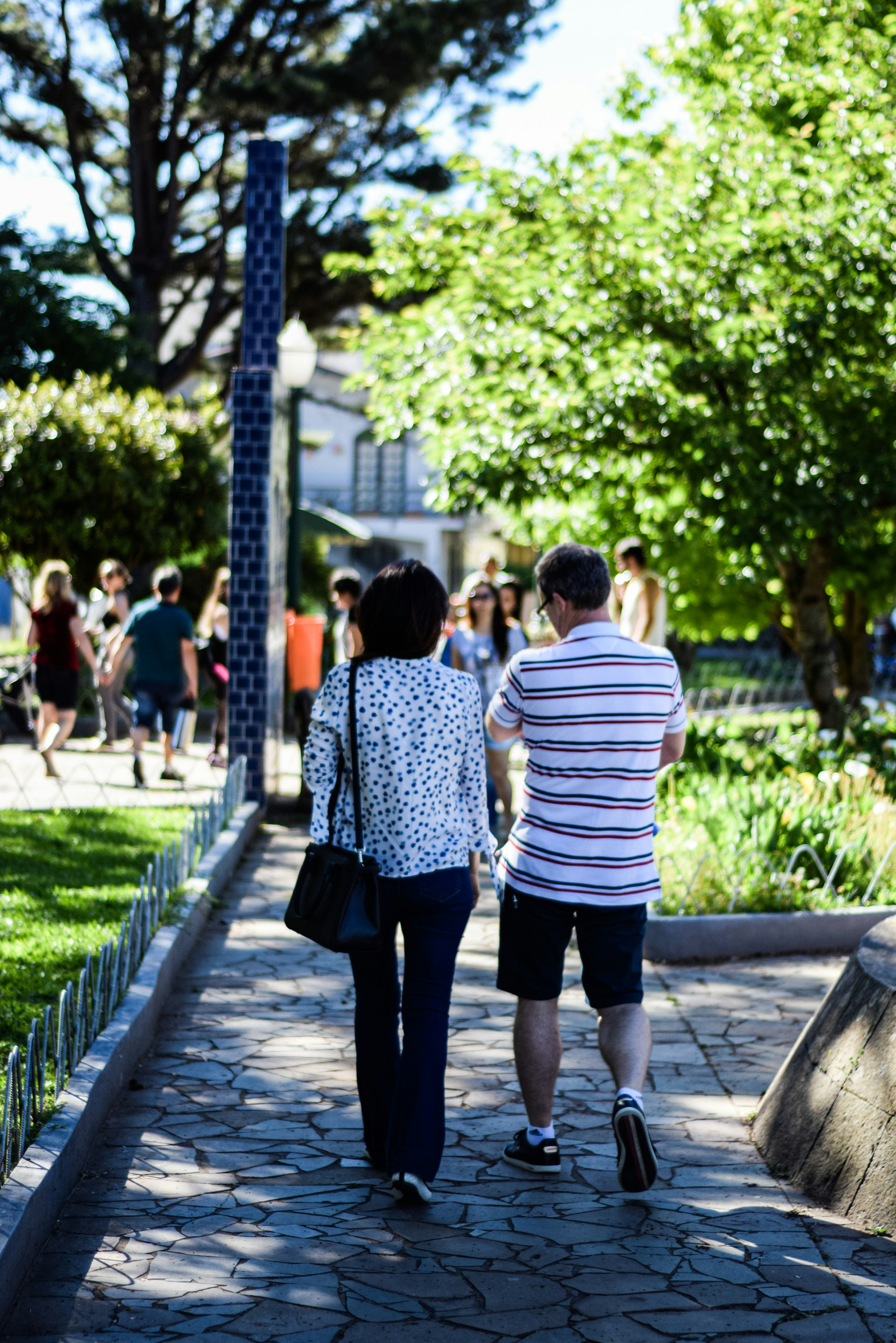 Couple walking hand in hand along a stone pathway in a vibrant park, surrounded by lush greenery and cheerful visitors.