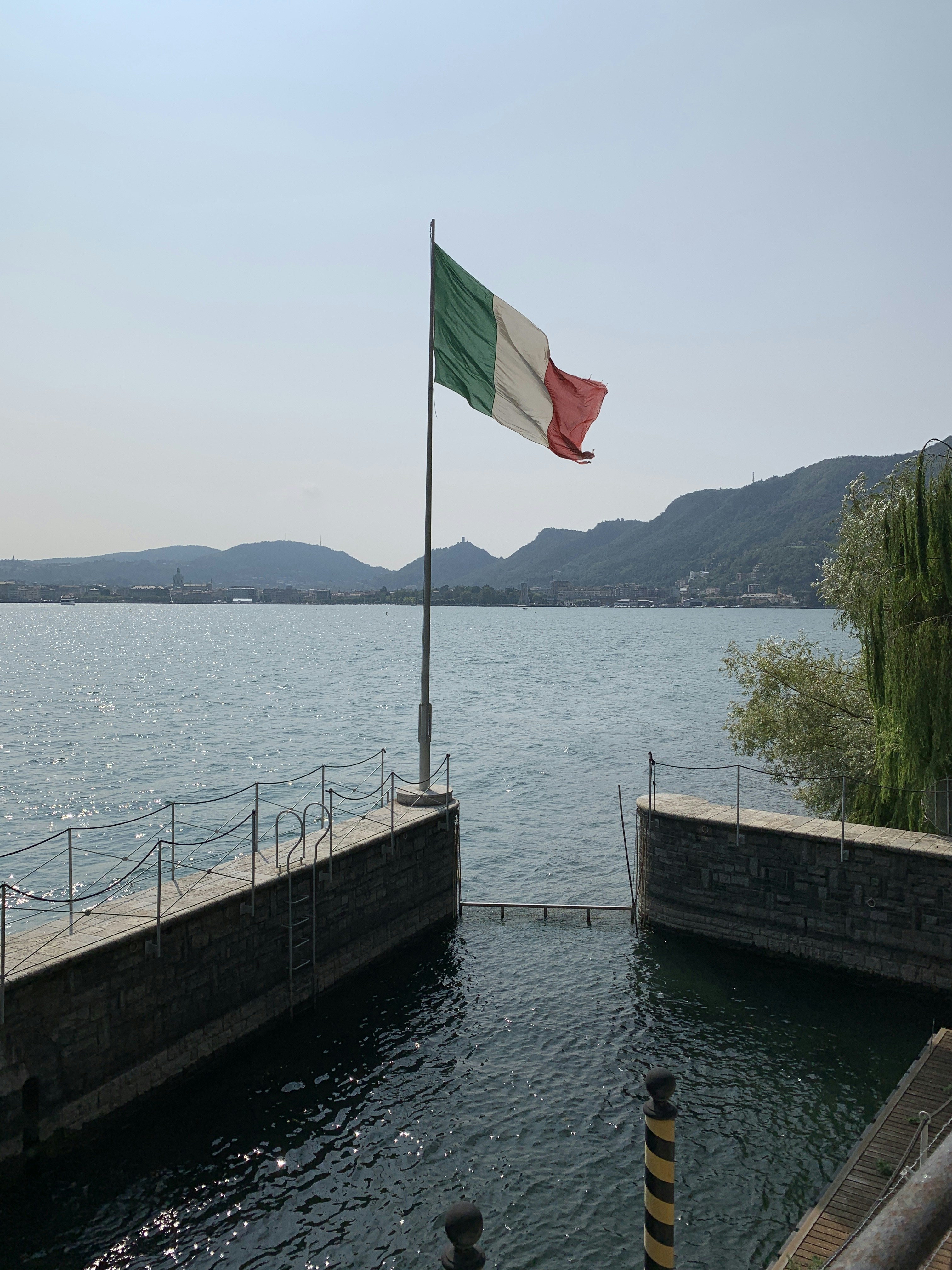 Italian flag waving proudly above a calm lake, with mountains in the background and a stone dock in the foreground.