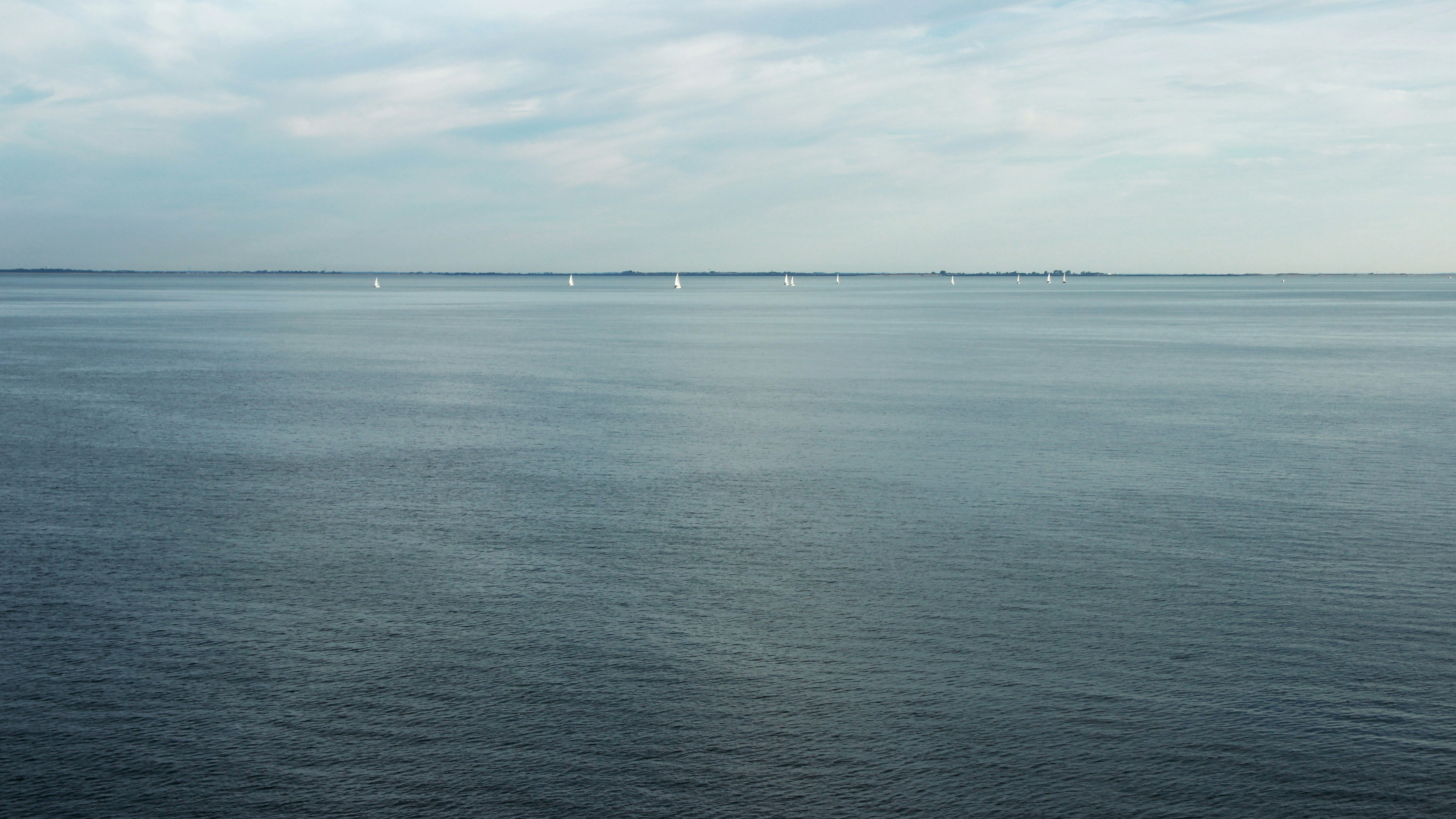 Calm sea with distant sailboats gliding across the water under a soft, cloudy sky.