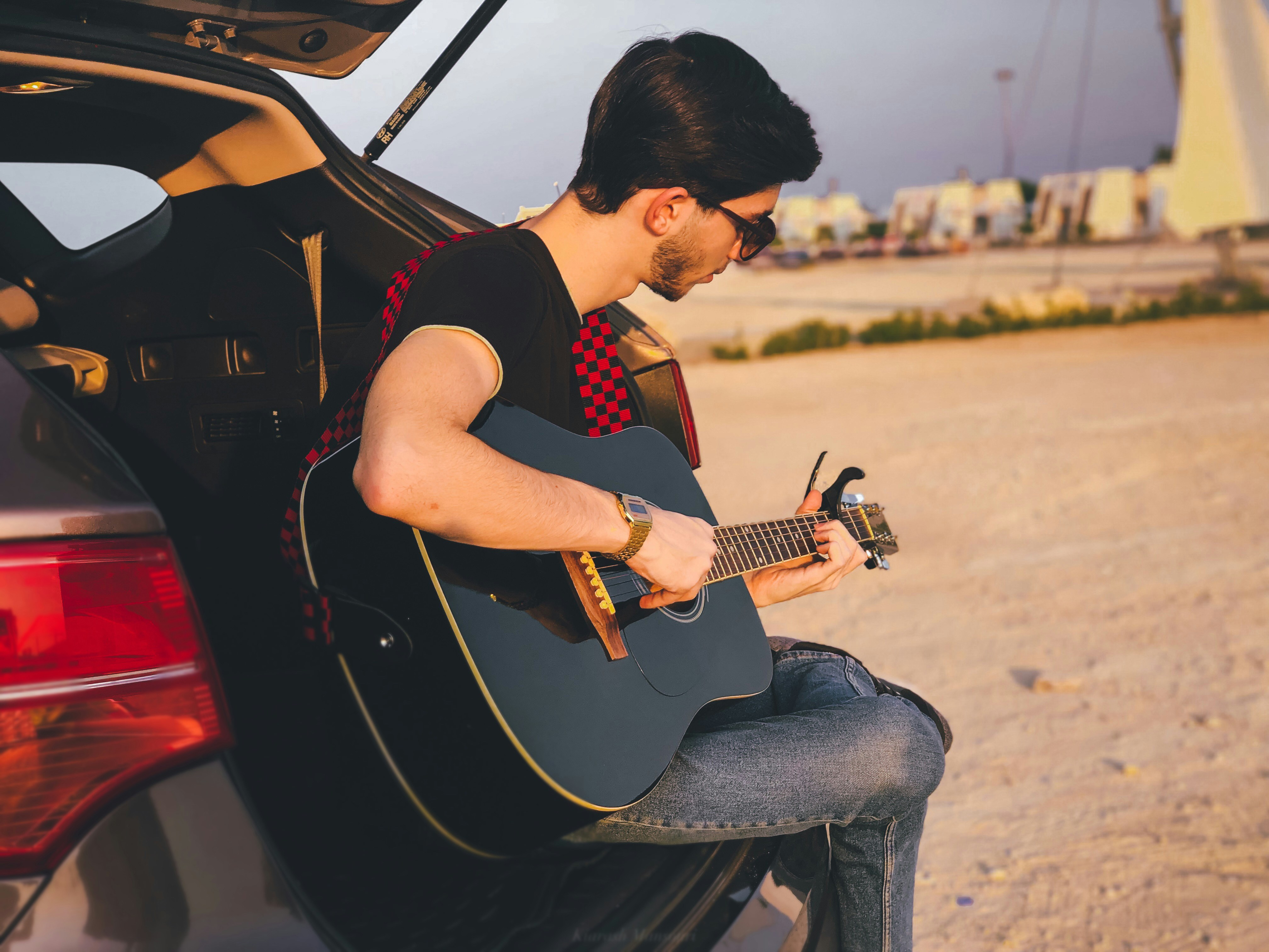 Young man playing acoustic guitar while seated at the back of a car in a sandy outdoor setting.