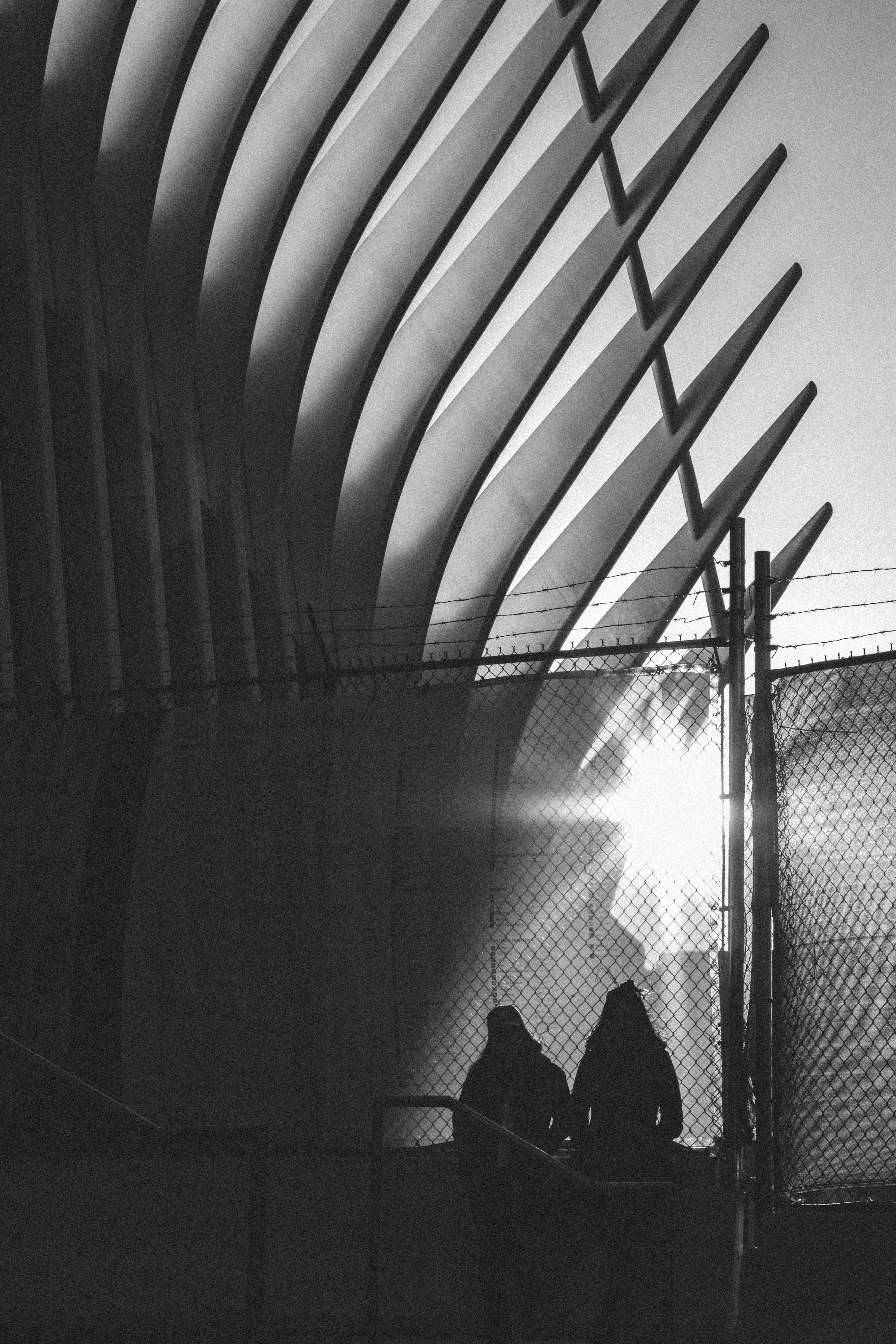 Two figures stand silhouetted against a striking modern structure, with sunlight piercing through a chain-link fence. The scene captures a moment of quiet reflection in an urban environment.