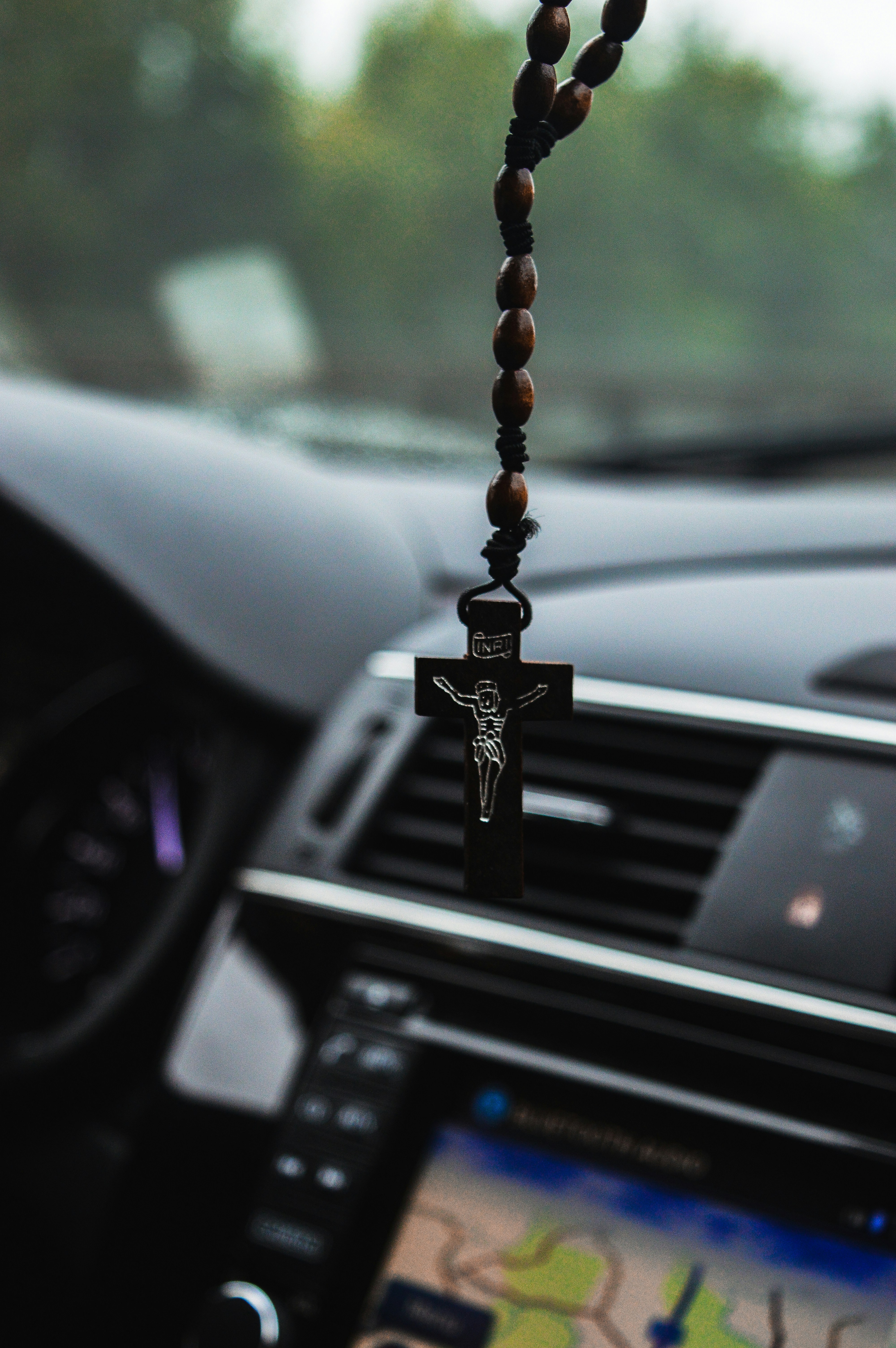 A wooden crucifix hangs from the rearview mirror, set against the backdrop of a car's dashboard with a navigation screen.