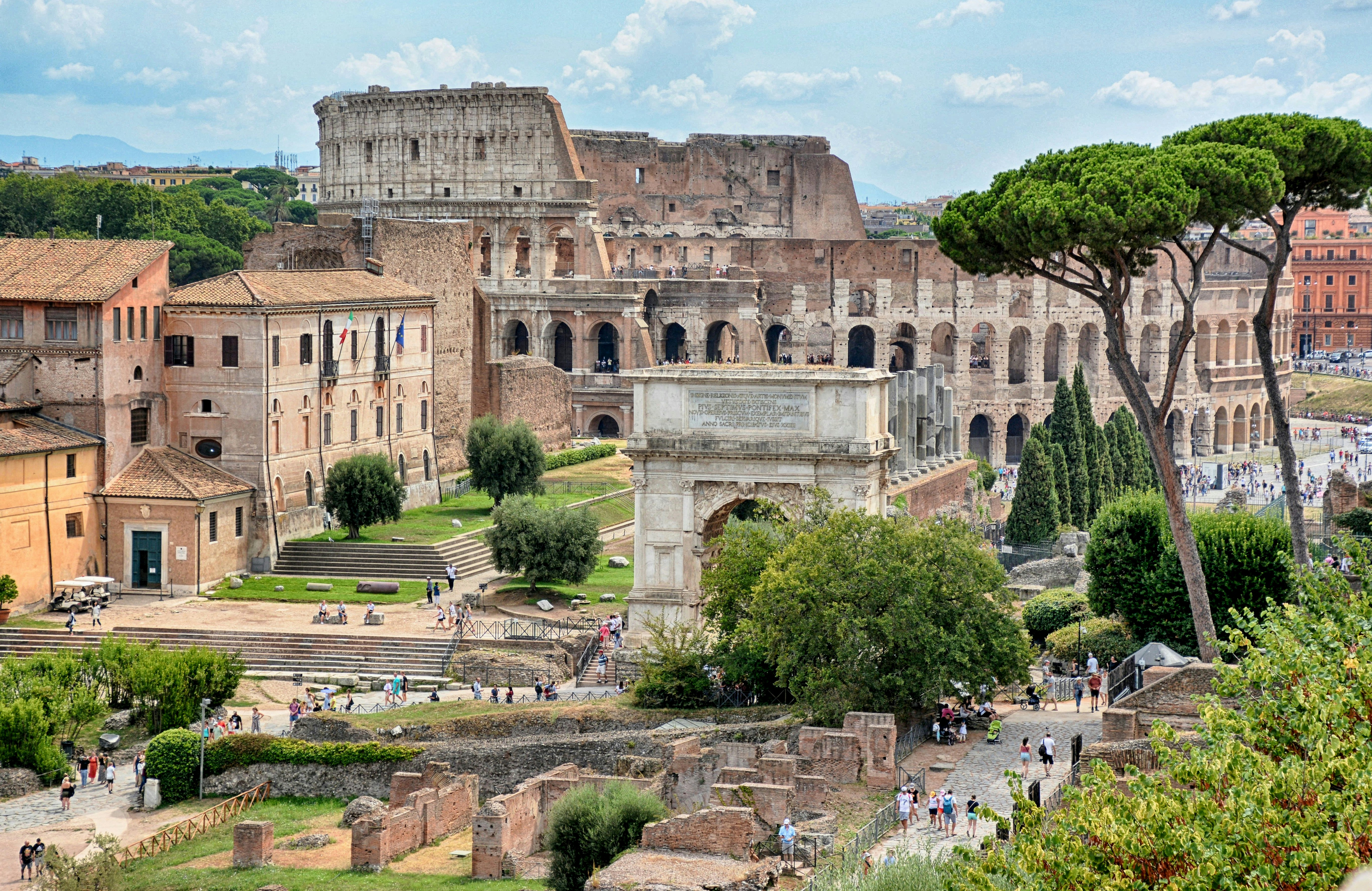 Aerial view of the Colosseum surrounded by lush greenery and historical structures under a partly cloudy sky.