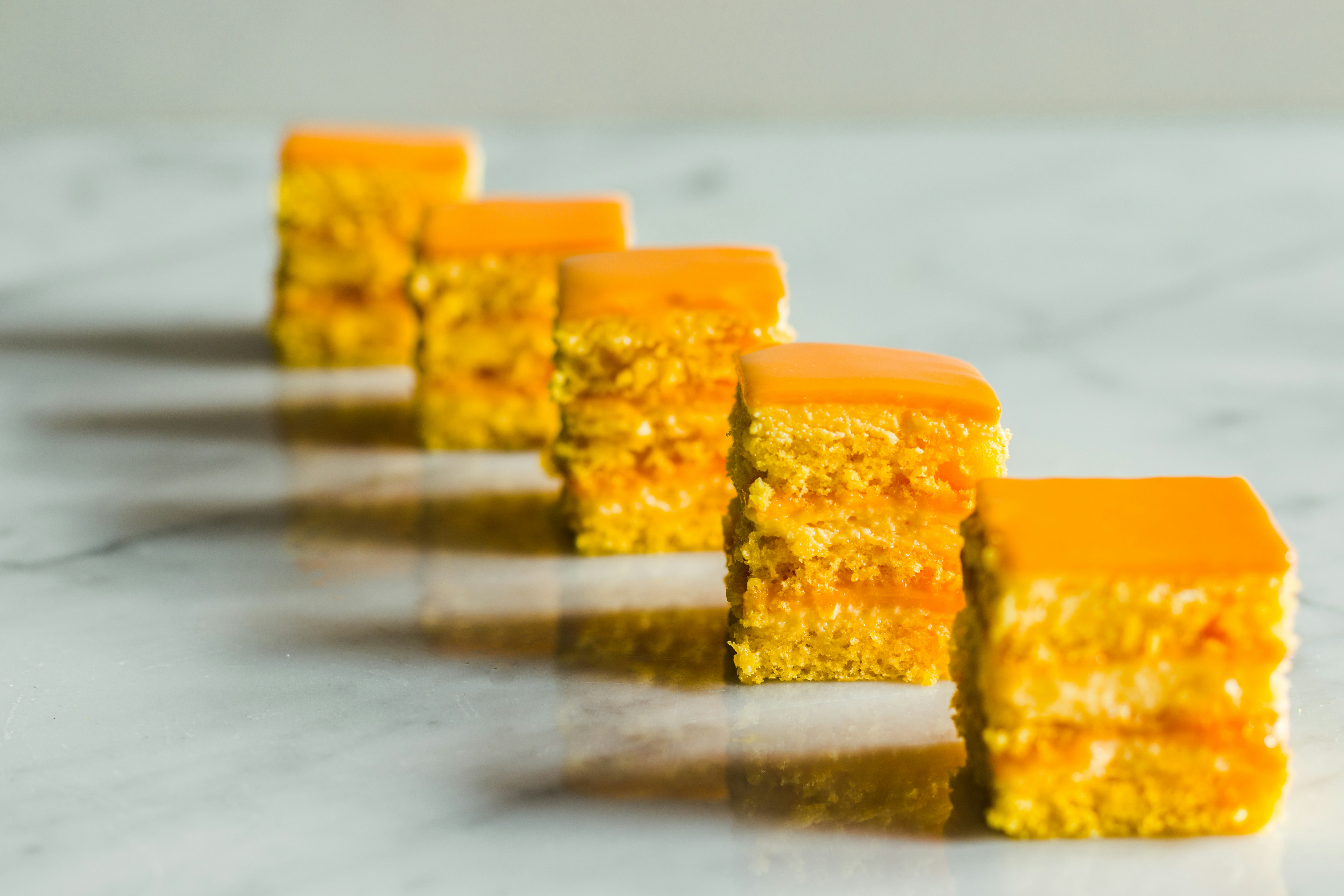 Five layers of yellow cake with glossy orange frosting arranged in a line on a marble surface.