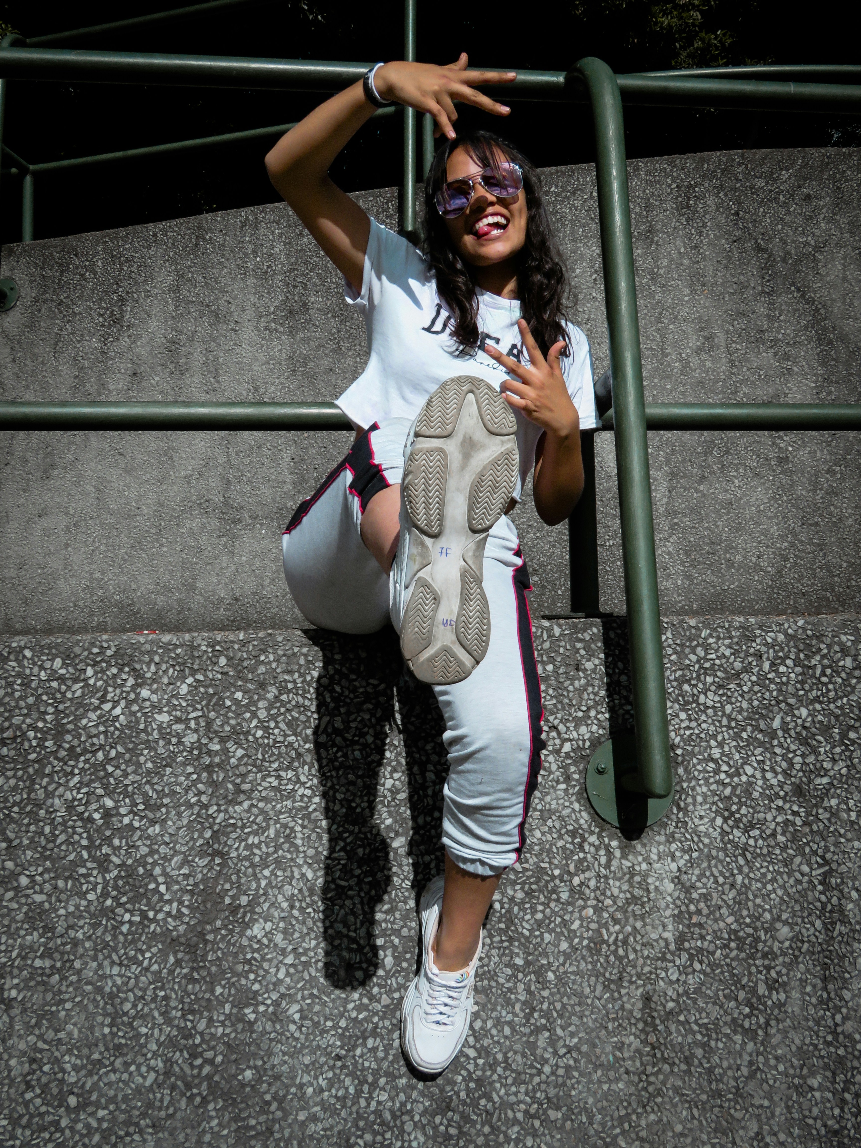 A young woman playfully poses with her foot raised, showcasing her sneaker while making a fun gesture. The vibrant urban backdrop adds to the lively atmosphere.