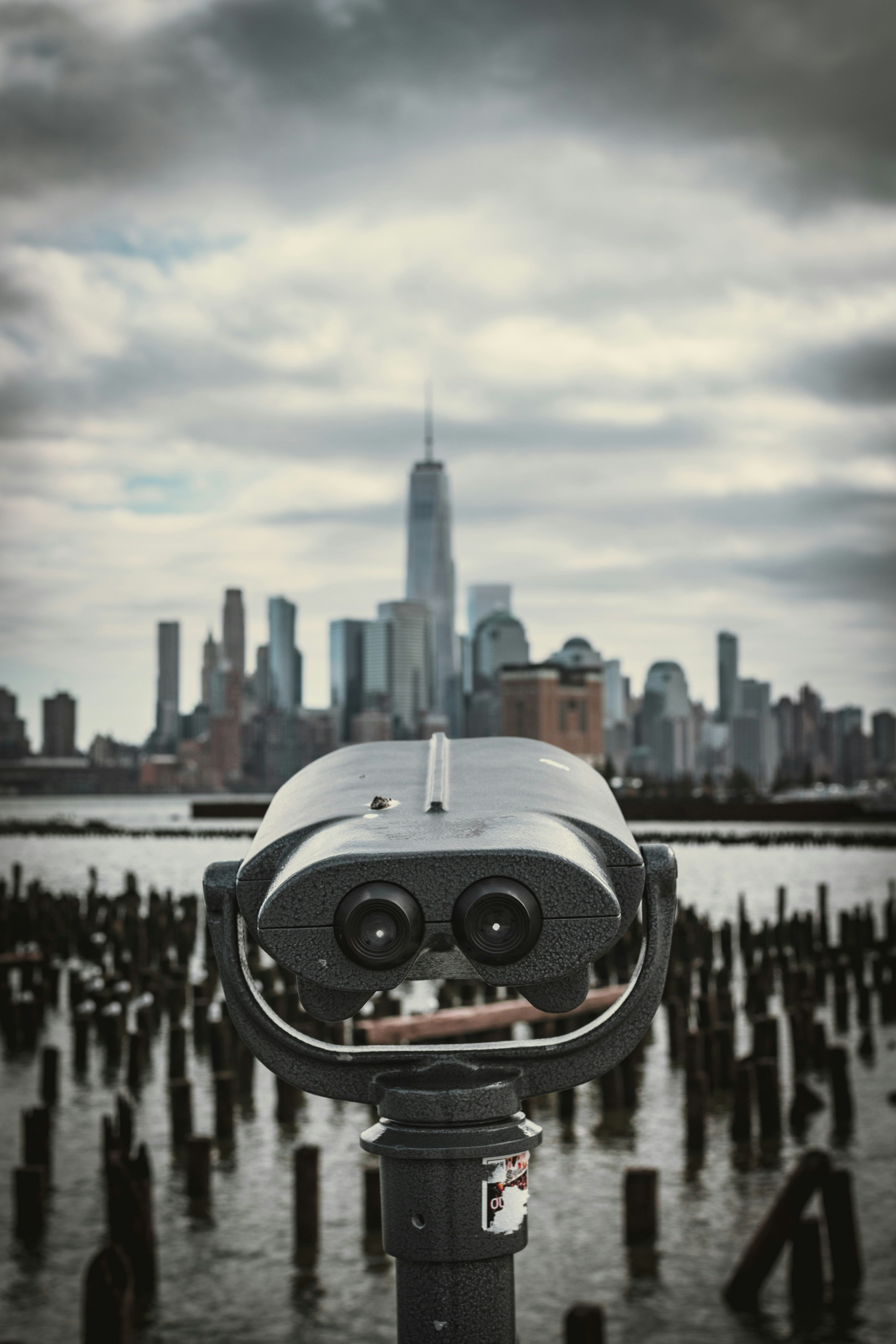 A vintage binocular viewer stands in the foreground, overlooking a city skyline marked by modern skyscrapers and weathered pilings in the water.