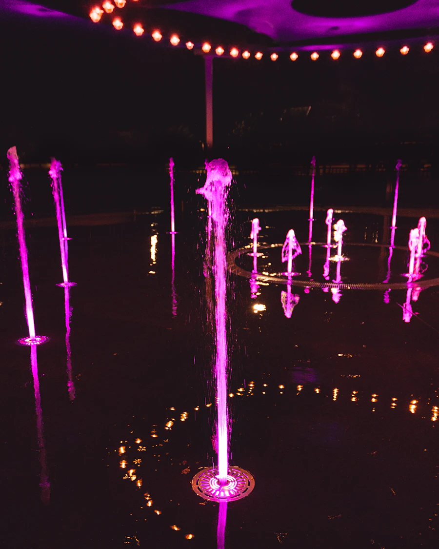 Several fountain jets glow bright magenta against a dark pool, with reflections and a ring of warm lights overhead. The tallest jet rises near the center foreground.