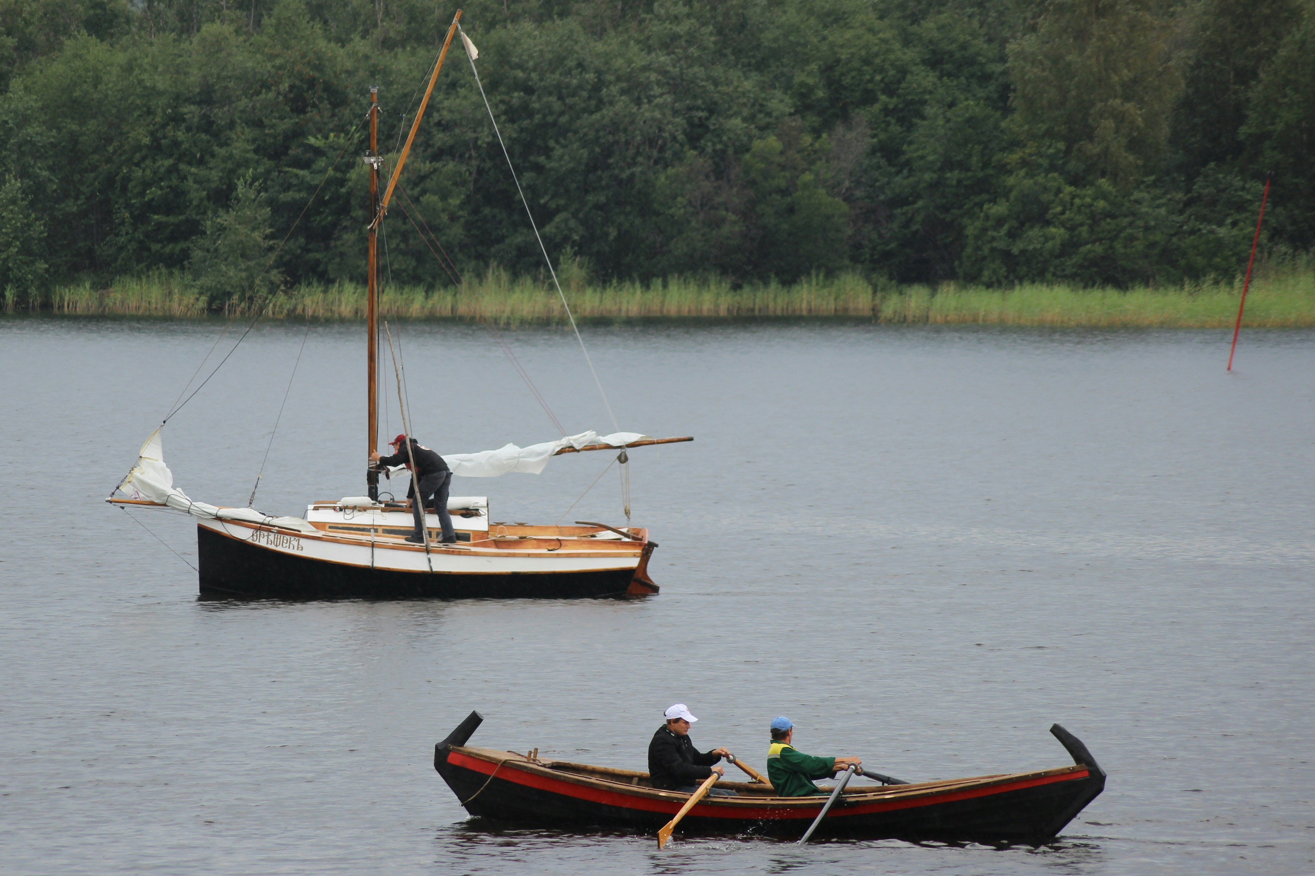 Two small boats drift on a calm lake: a white-sailed vessel ahead and two rowers in a dark hull foreground, with a tree-lined shoreline in the distance.