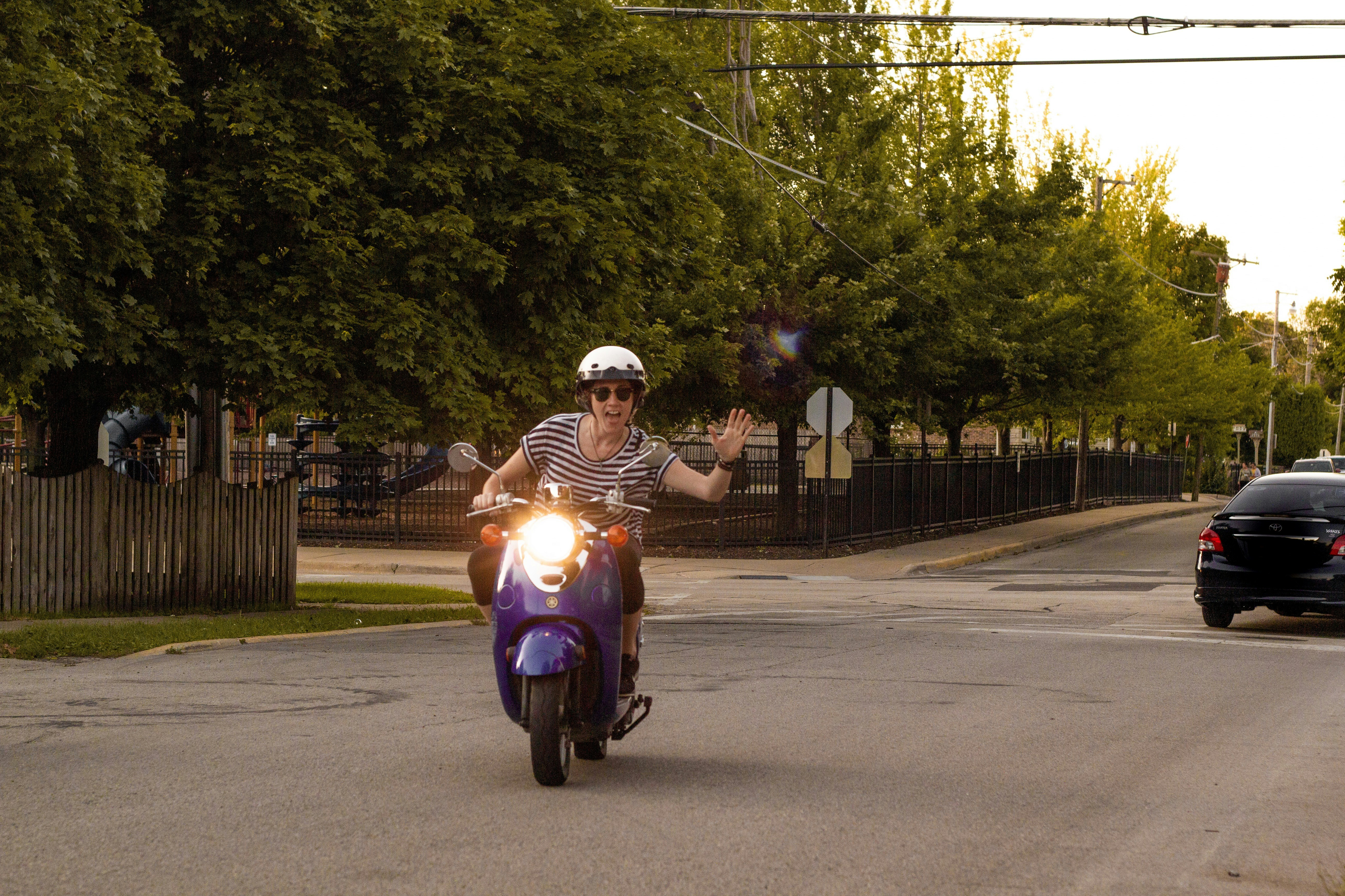 Person waving enthusiastically while riding a scooter down a tree-lined street.