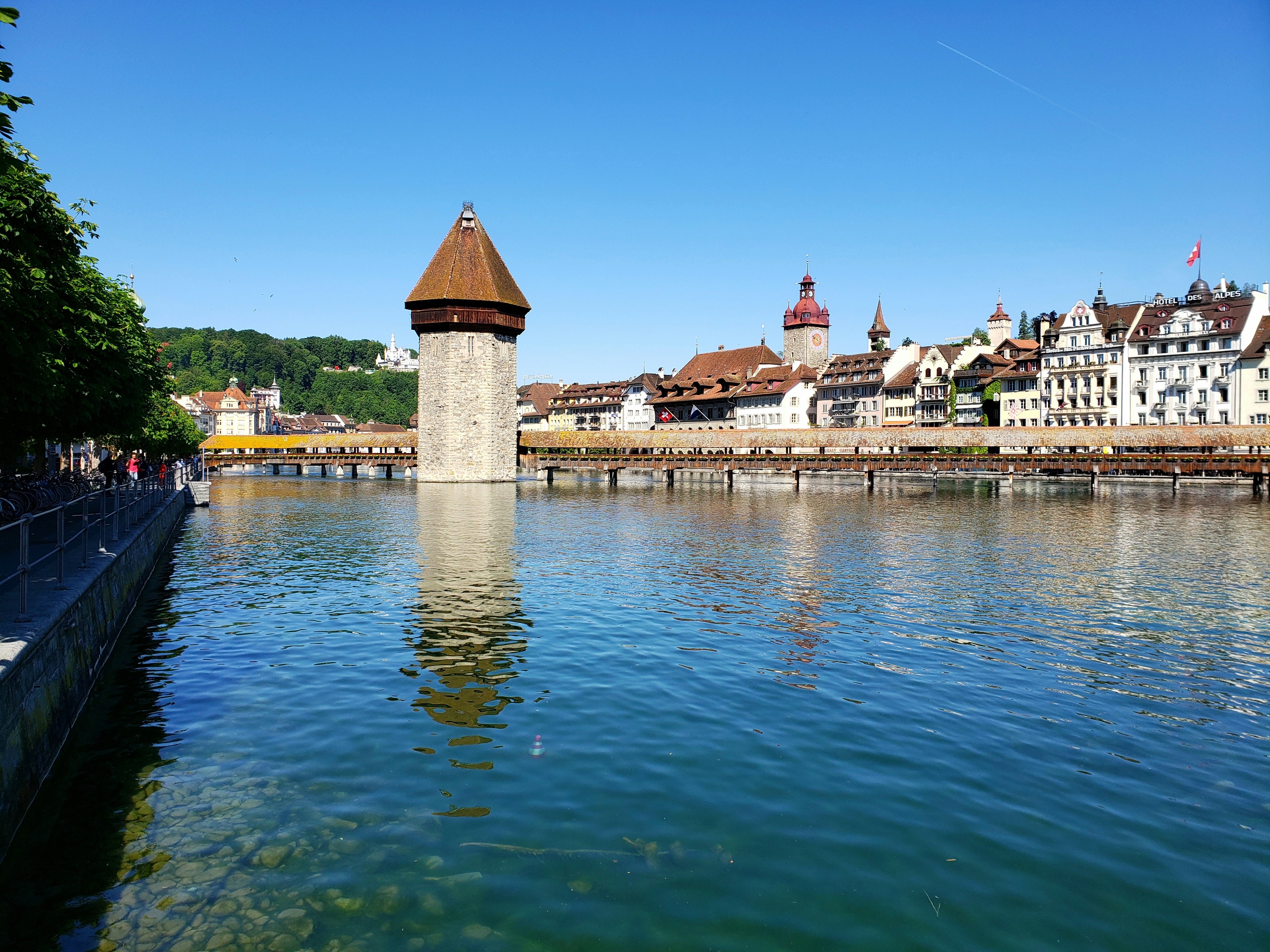 Medieval tower stands reflected in the calm river, with a backdrop of historic buildings under a clear blue sky.