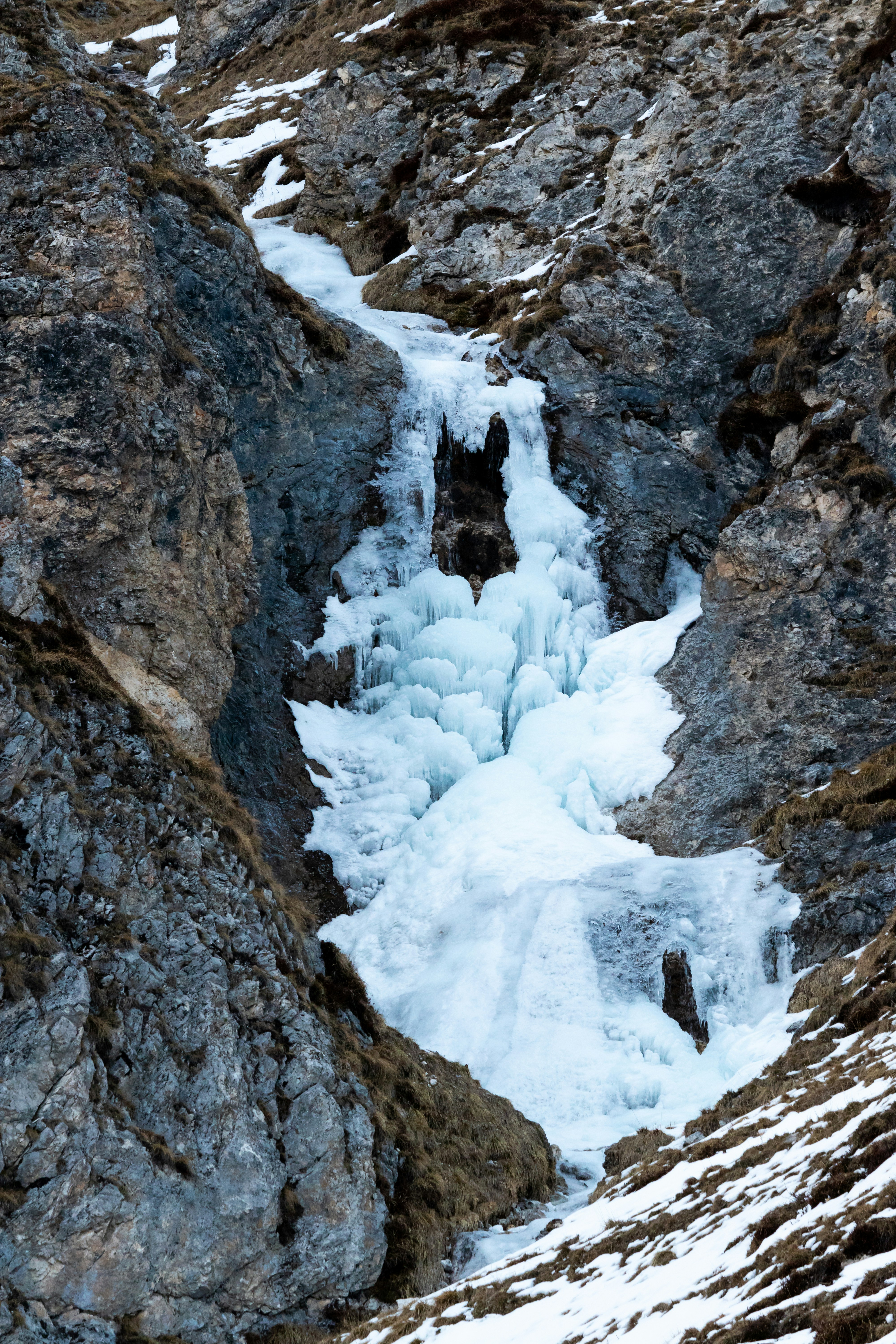 A frozen waterfall cascading down rocky terrain, surrounded by rugged mountain landscape and patches of snow.