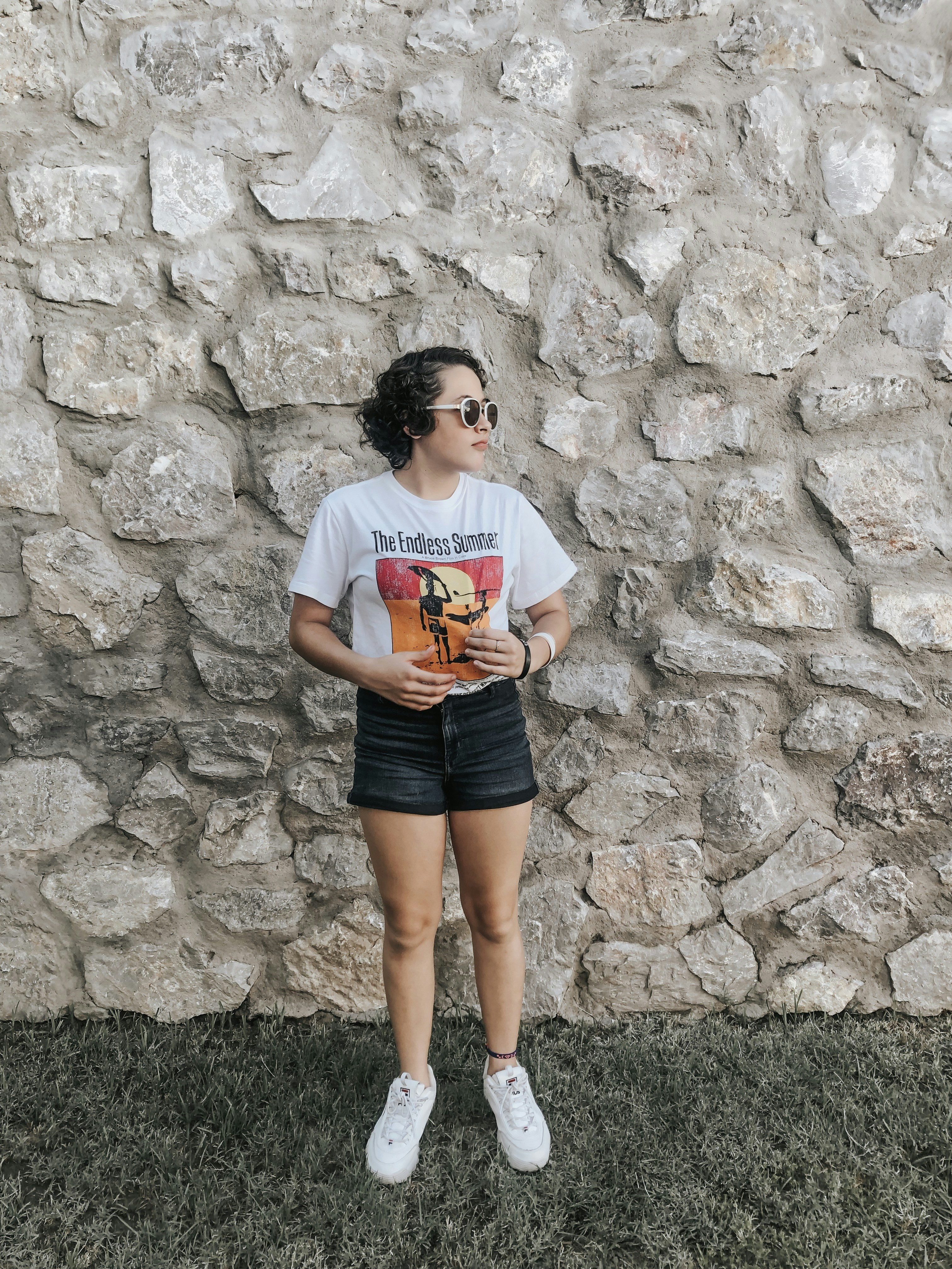 Individual posing in casual attire against a textured stone wall, exuding a relaxed summer aesthetic.
