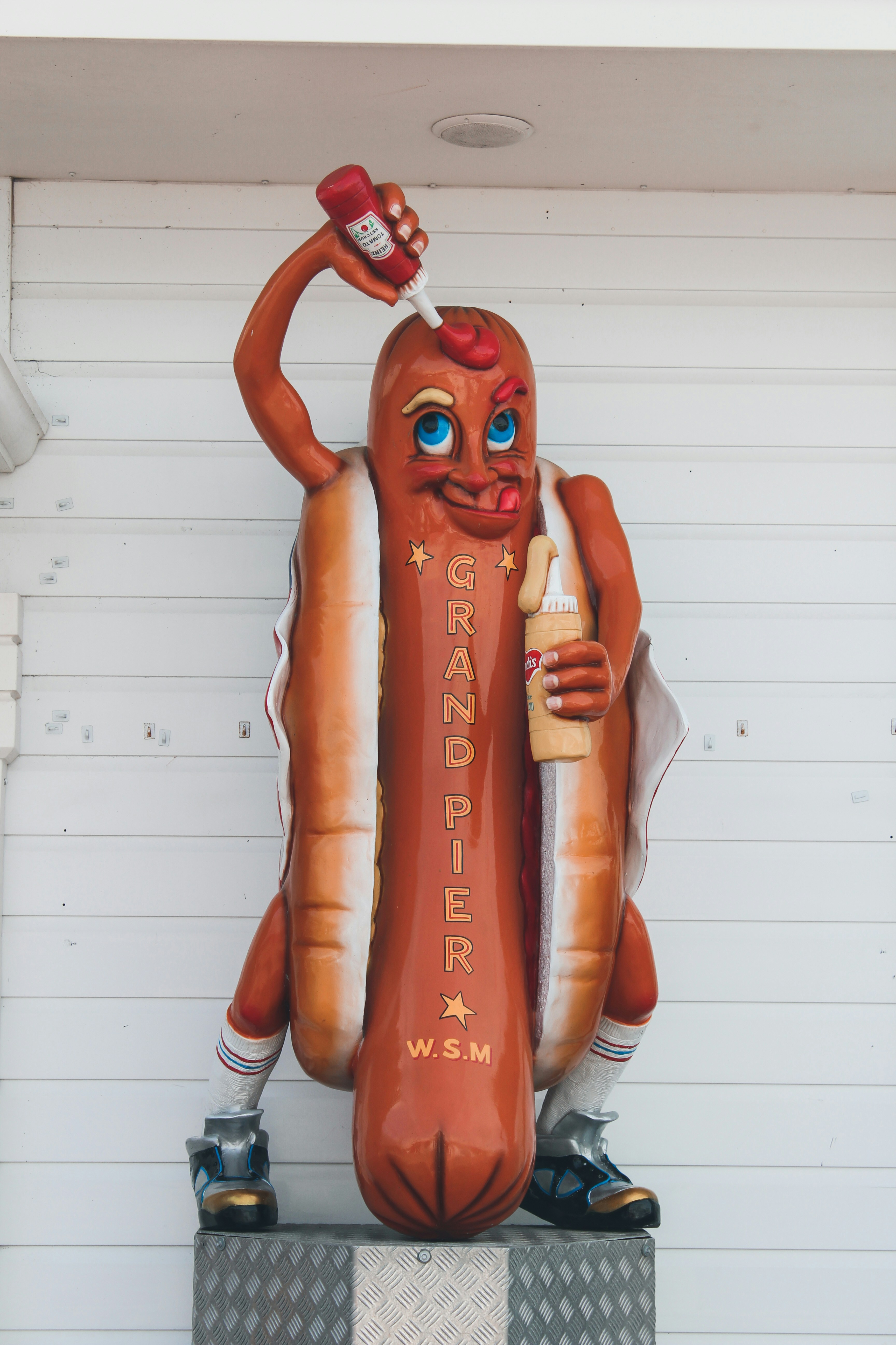 A whimsical hot dog mascot playfully dispensing ketchup while holding a drink, promoting the Grand Pier. The vibrant colors and playful design invite attention.