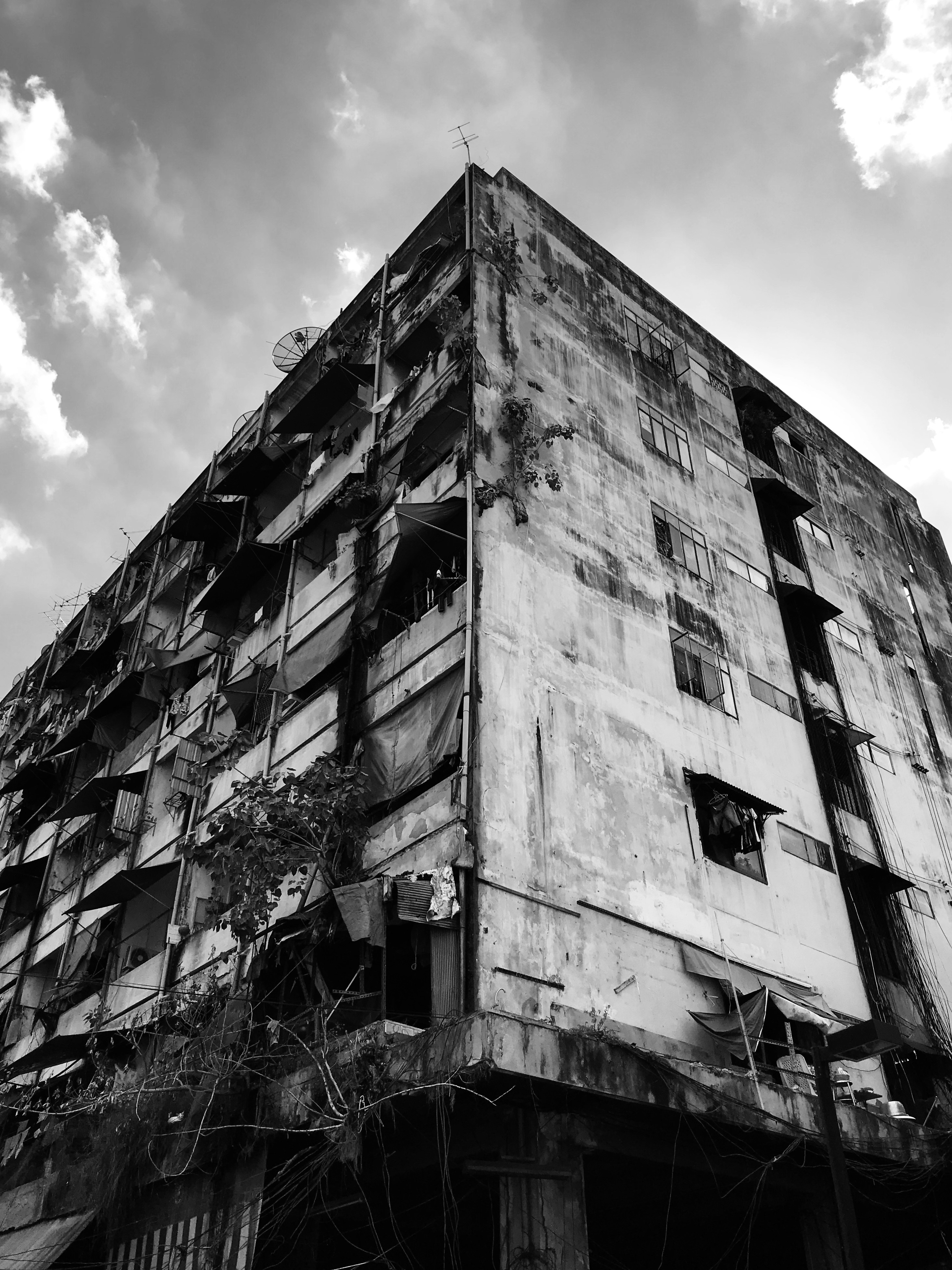 A weathered high-rise building stands in stark contrast against a dramatic sky, showcasing peeling paint and overgrown vegetation.