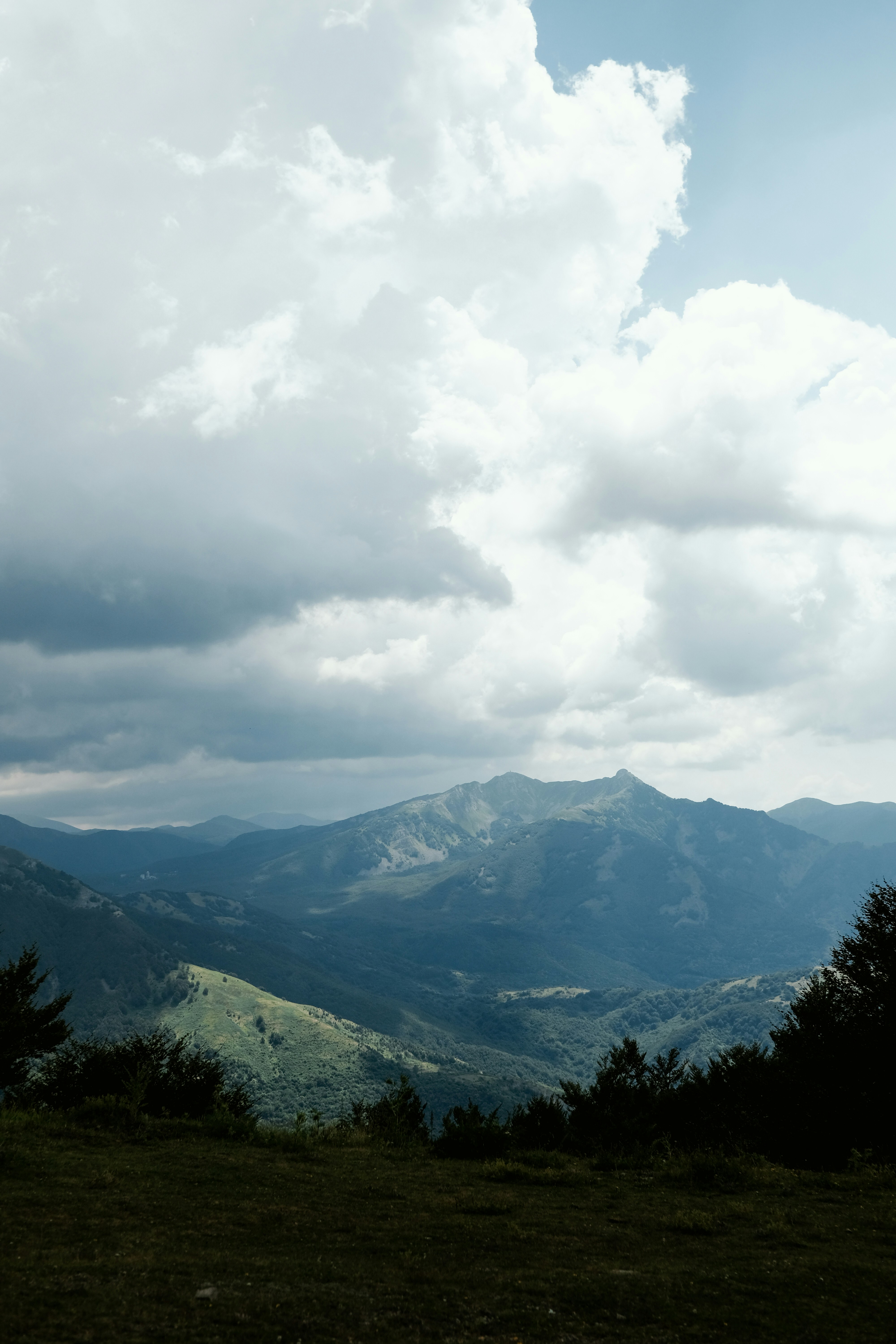 Dramatic mountain landscape under a cloudy sky, showcasing rolling hills and distant peaks. The scene evokes a sense of tranquility and vastness.