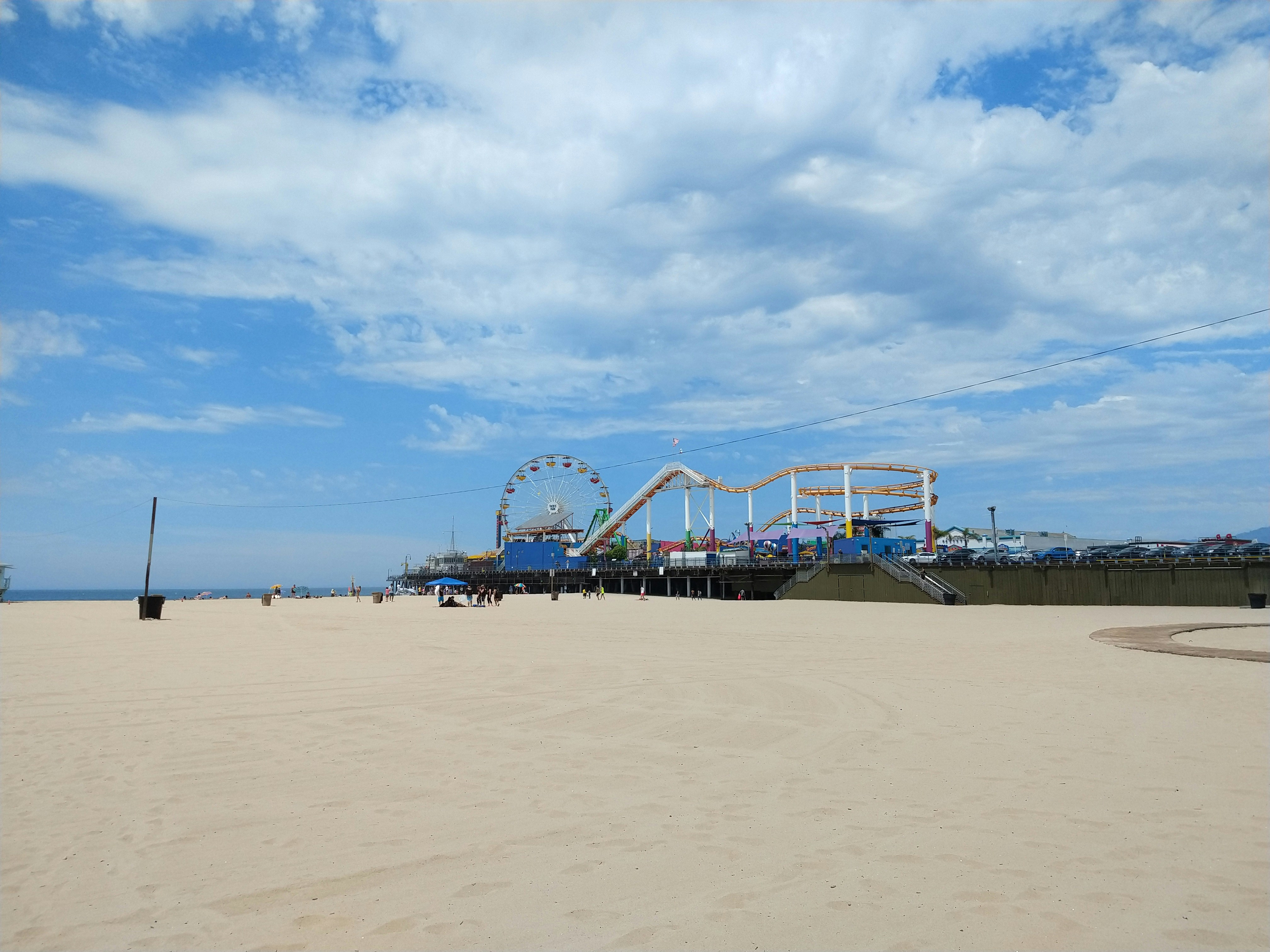 Amusement park rides and a Ferris wheel stand against a backdrop of clouds over a sandy beach. A lively scene captures the essence of summer fun.