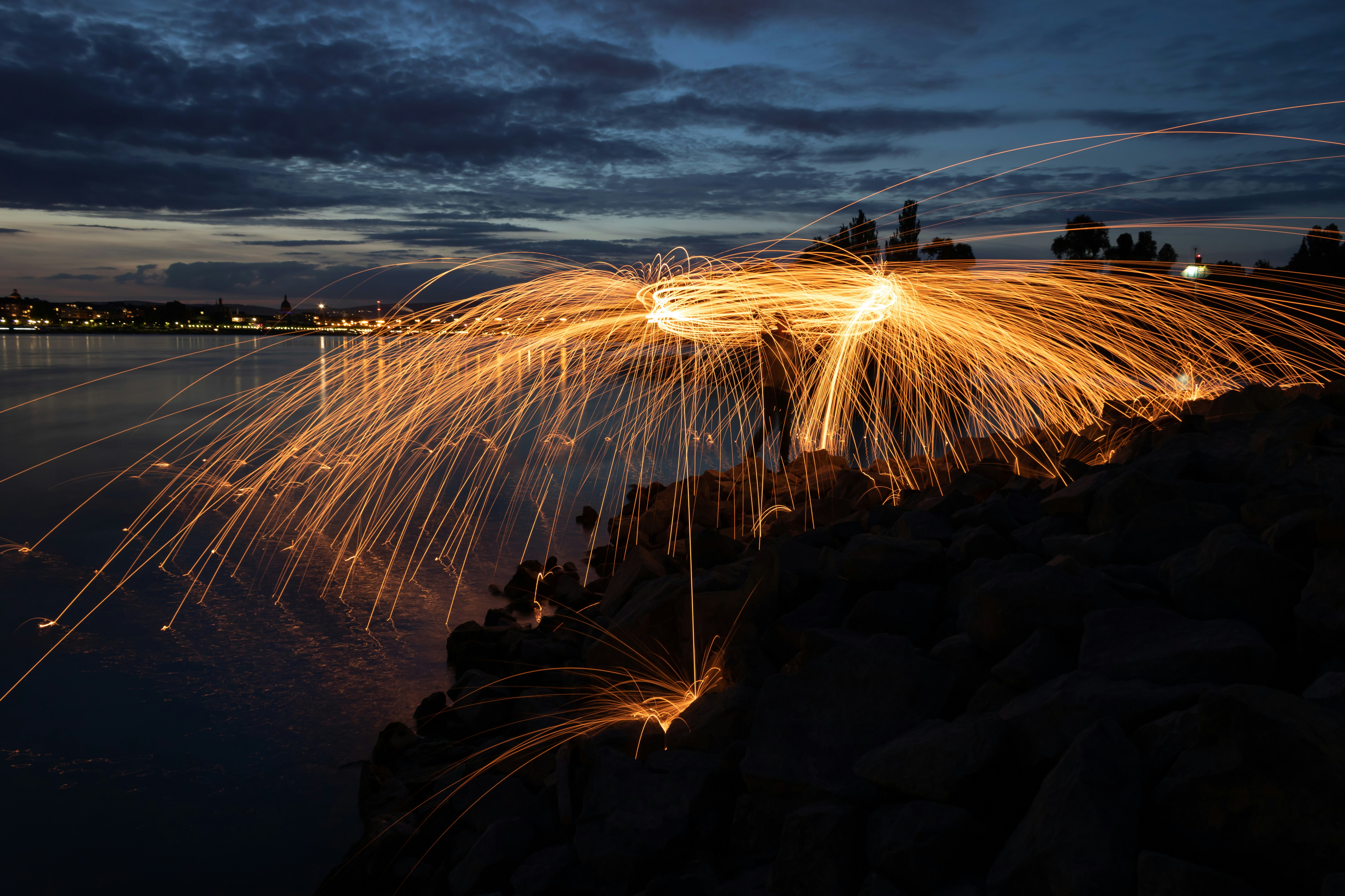 Long exposure of spinning steel wool creating fiery trails over a rocky shore at dusk.