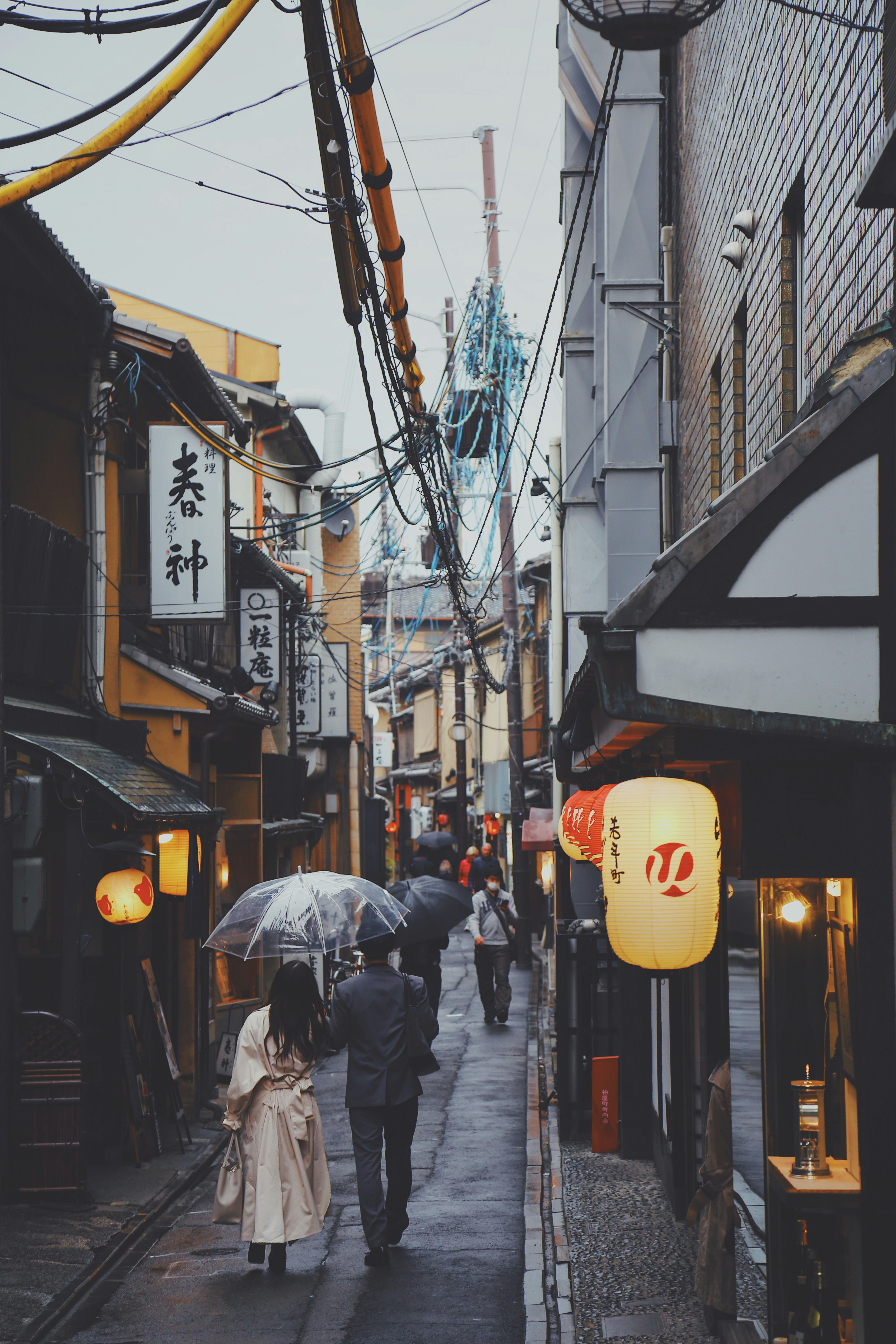 Couple walking hand-in-hand under an umbrella in a narrow street adorned with traditional lanterns and vintage signs.